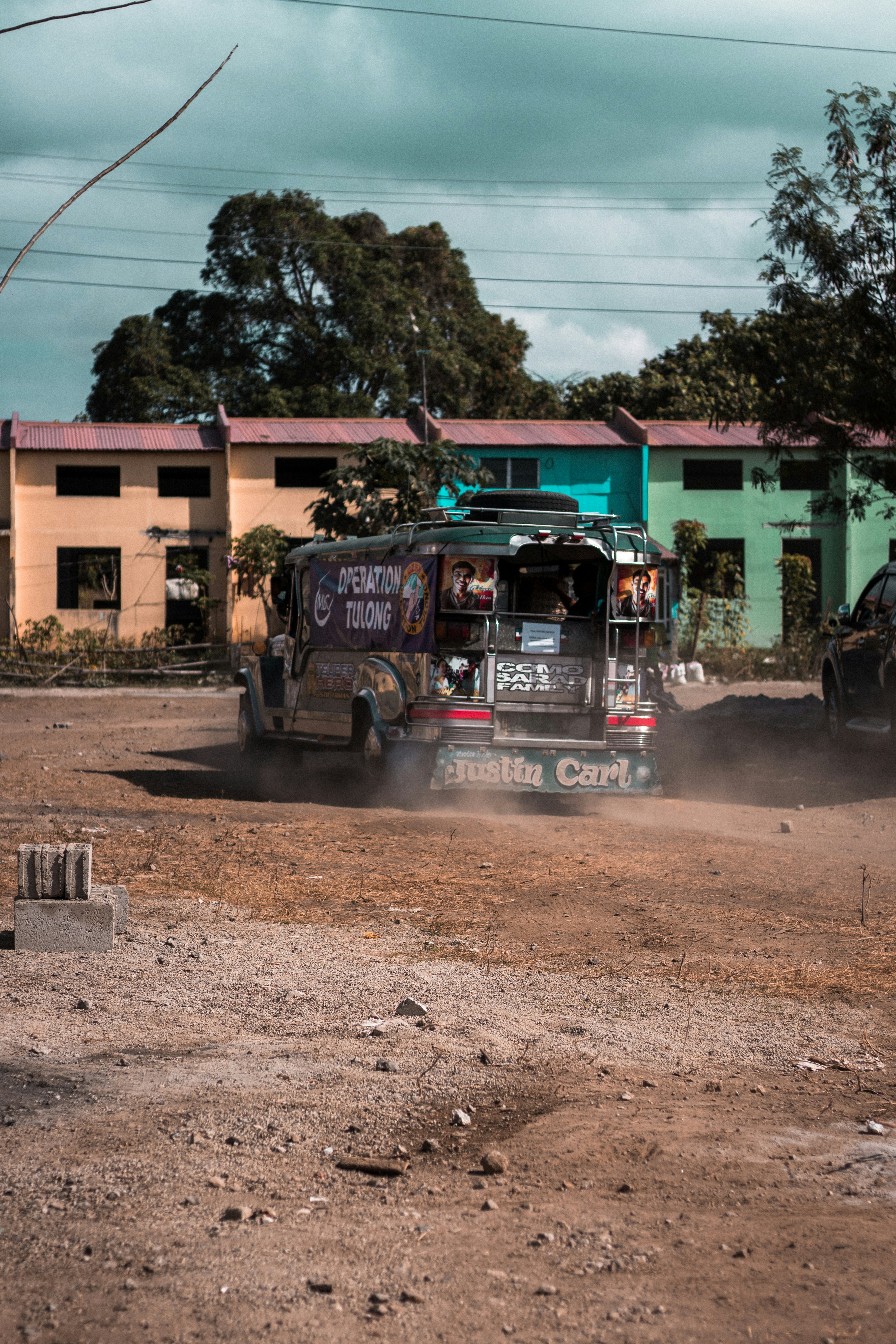 A colorful jeepney kicking up dust as it navigates a rural landscape, flanked by vibrant buildings in the background.