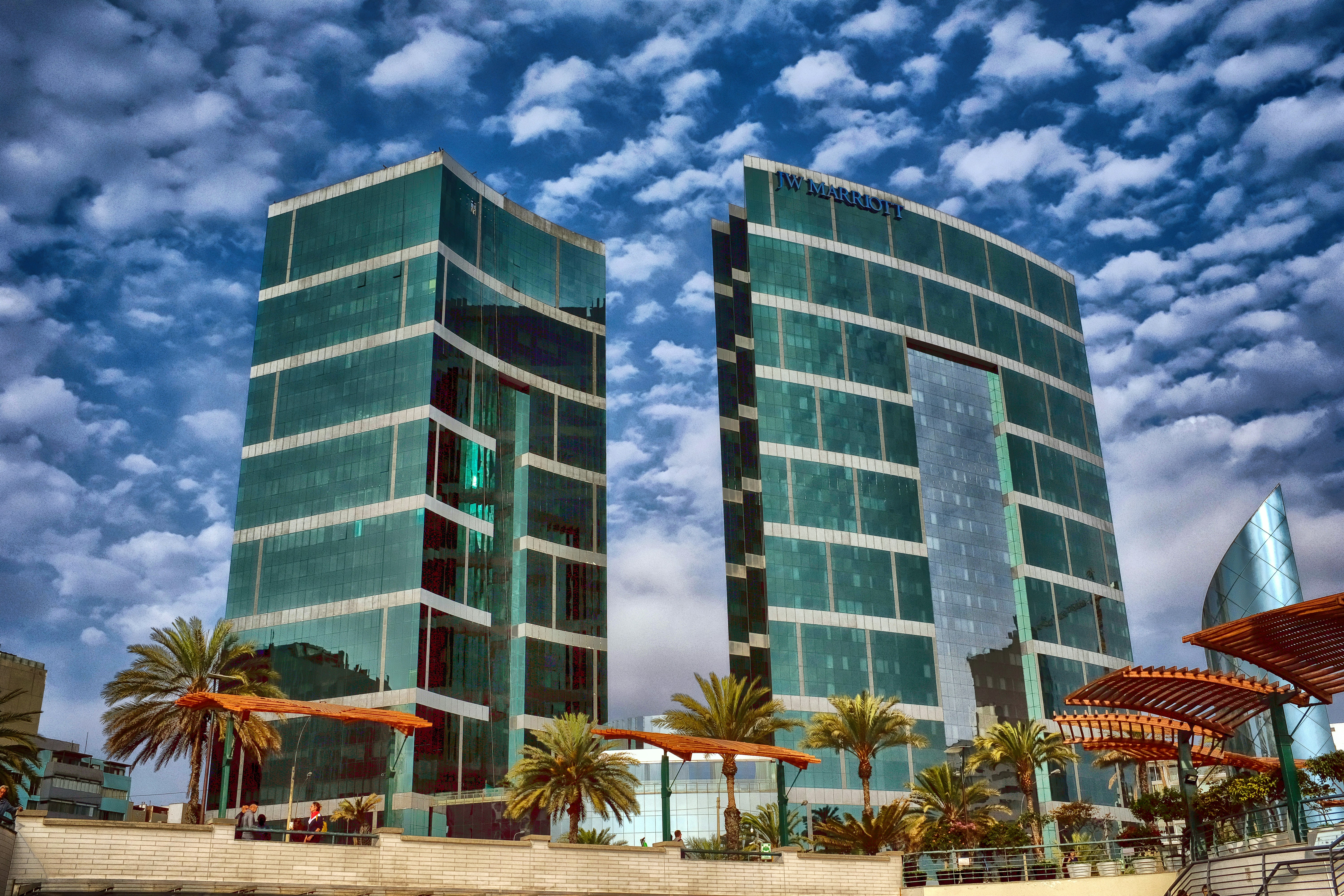 Modern glass buildings framed by palm trees under a sky filled with textured clouds.