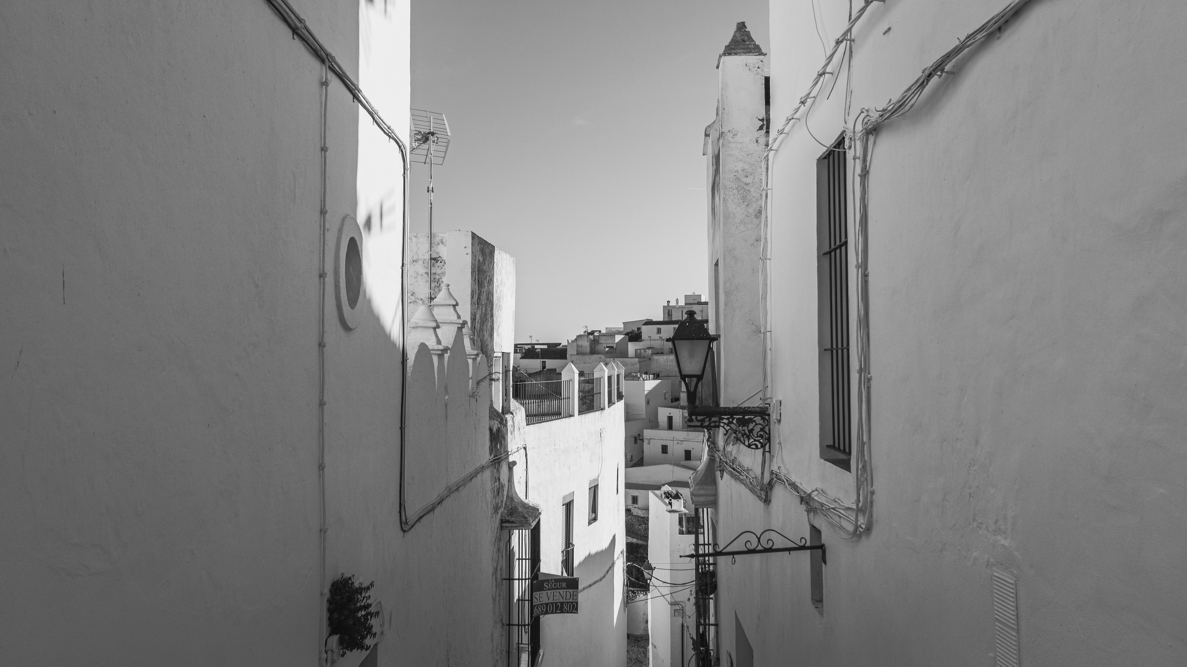 Narrow alleyway flanked by whitewashed buildings, leading to a distant view of rooftops under a clear sky.