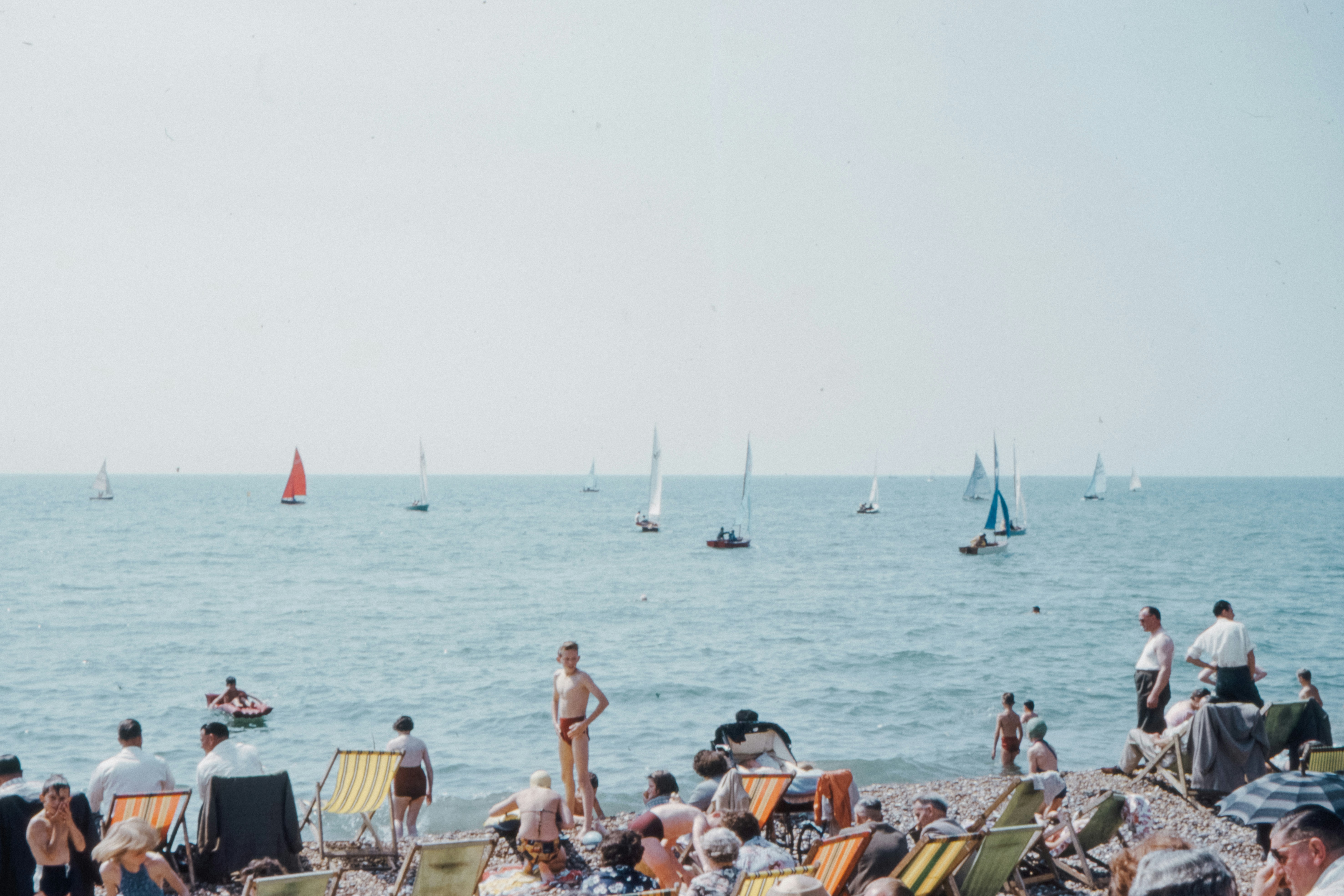 Crowded beach scene with people relaxing on deck chairs, overlooking a sea dotted with sailboats under a clear sky.