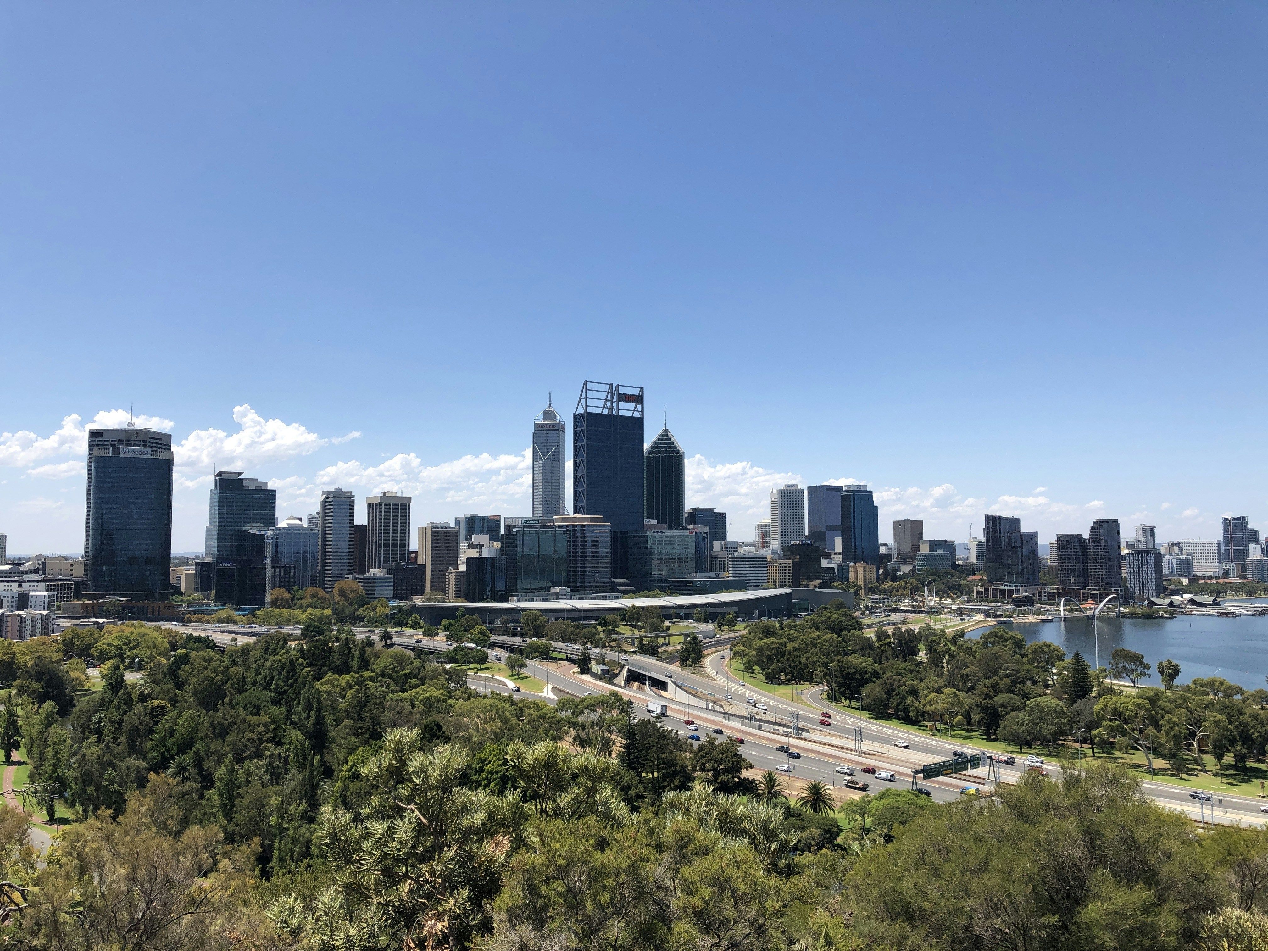 City skyline with lush greenery and a winding highway under a clear blue sky.