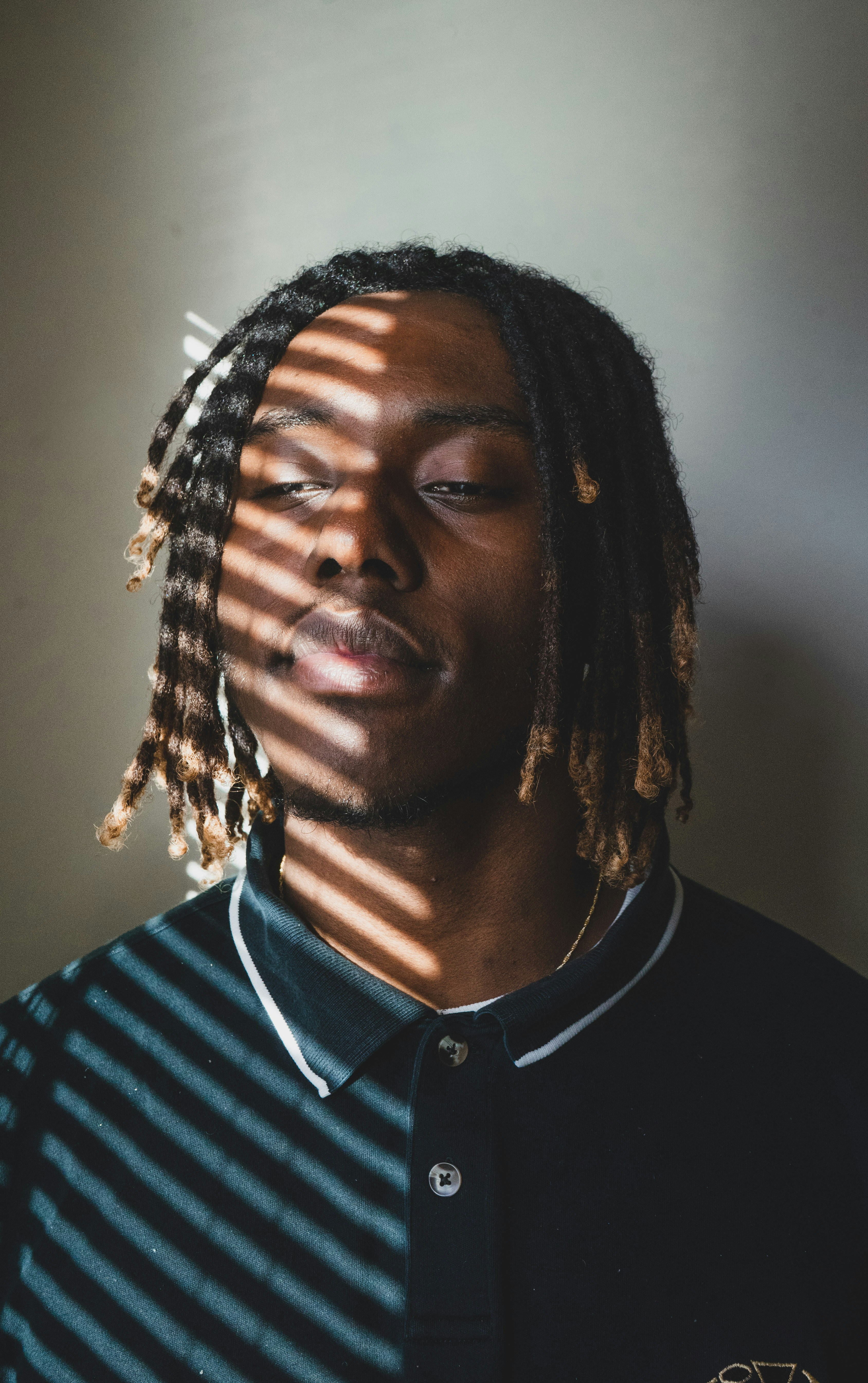 A young man with dreadlocks is illuminated by soft light filtering through striped shadows, creating an intriguing interplay of light and texture.