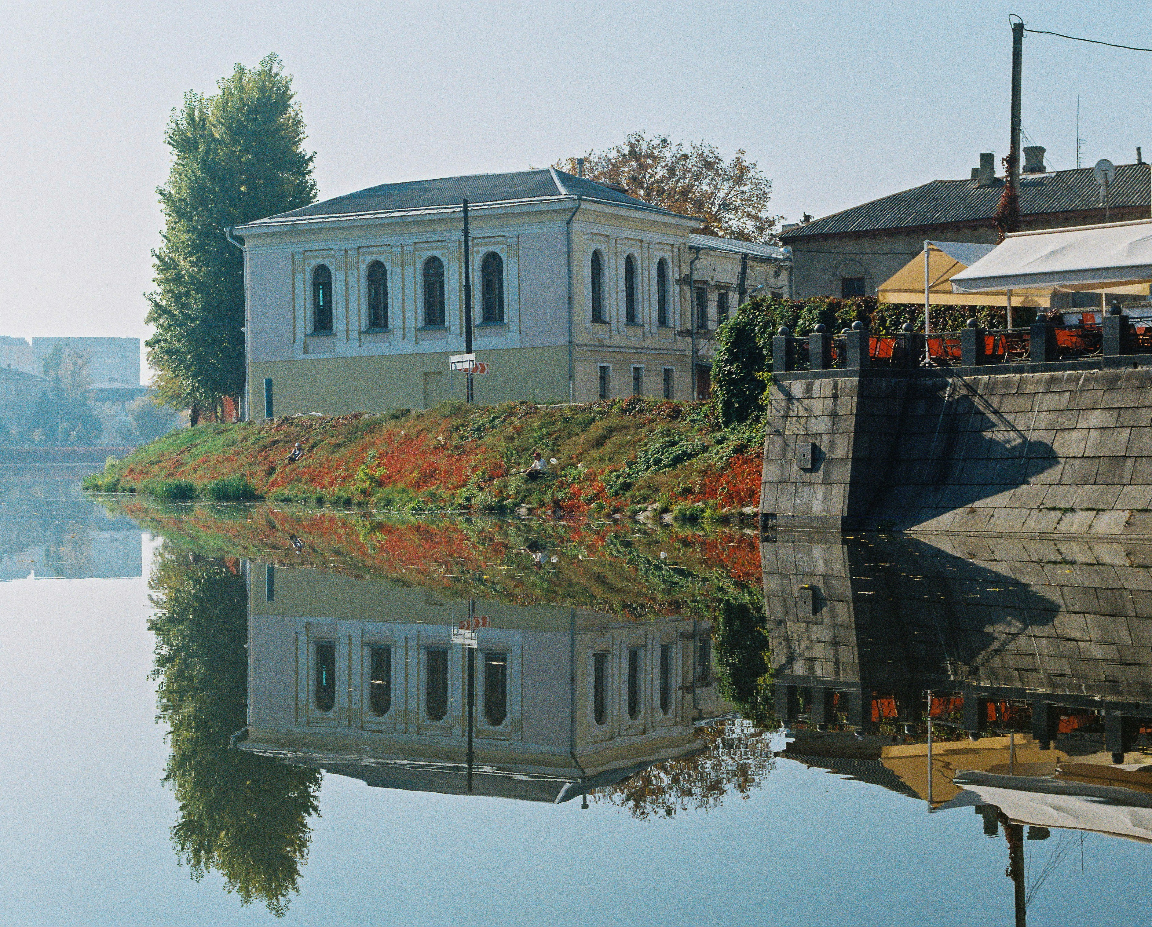 Photograph of a quiet riverside with a whitewashed building and its mirrored reflection in the calm water.
