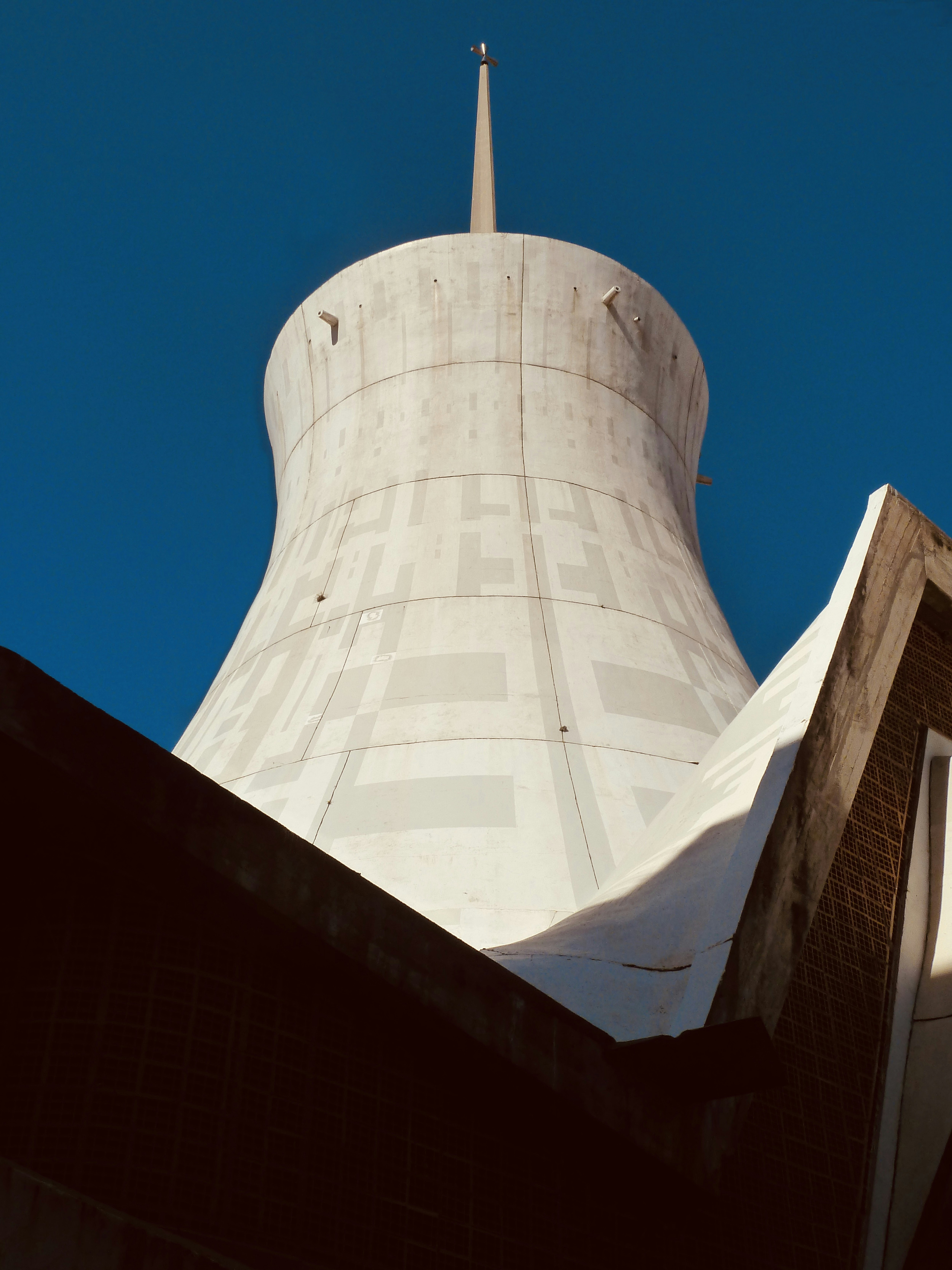 White cooling tower rises against a deep blue sky, framed by angular foreground rooftops.