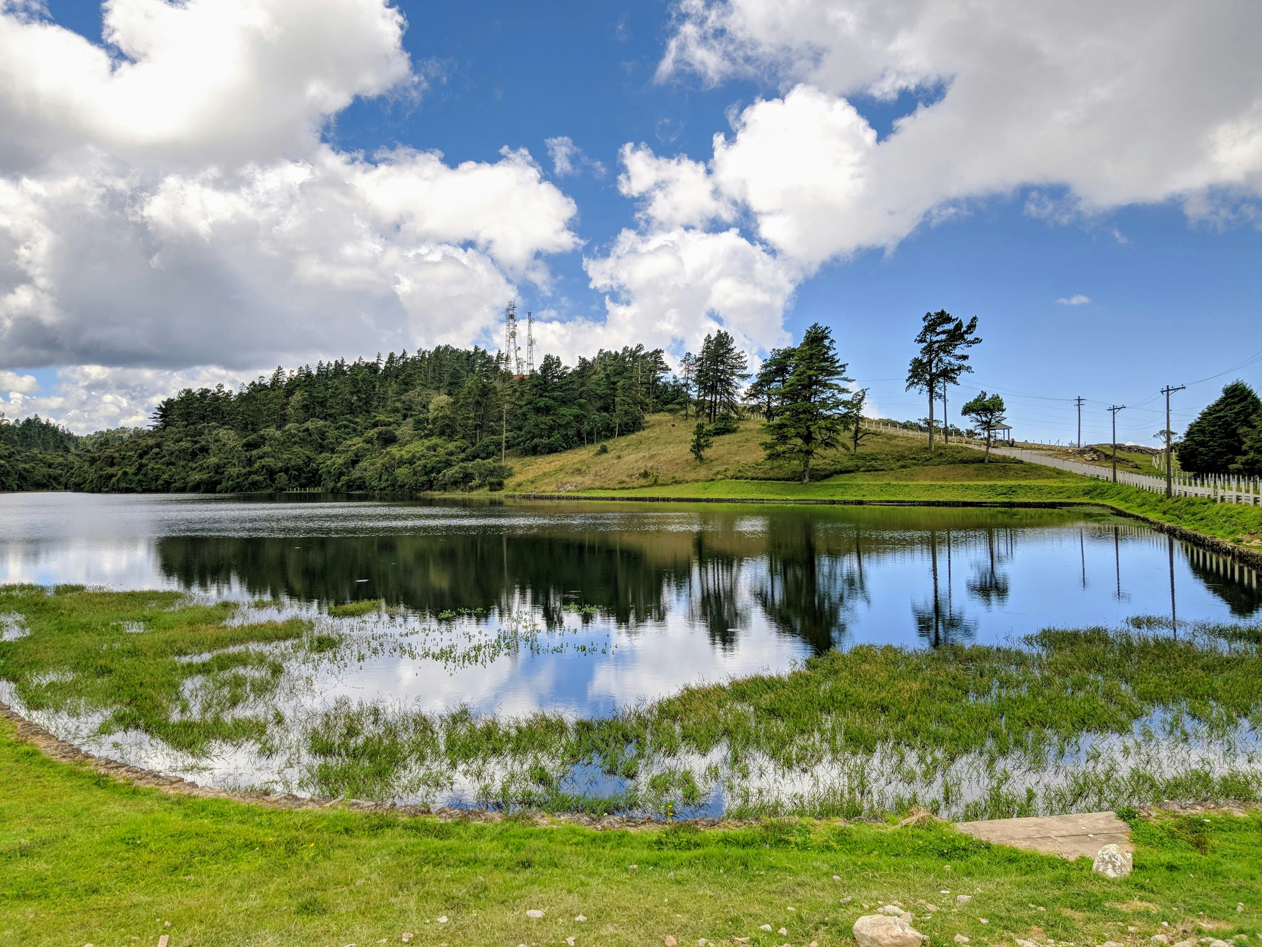 Clear lake reflecting lush green hills and scattered trees under a sky filled with fluffy white clouds.
