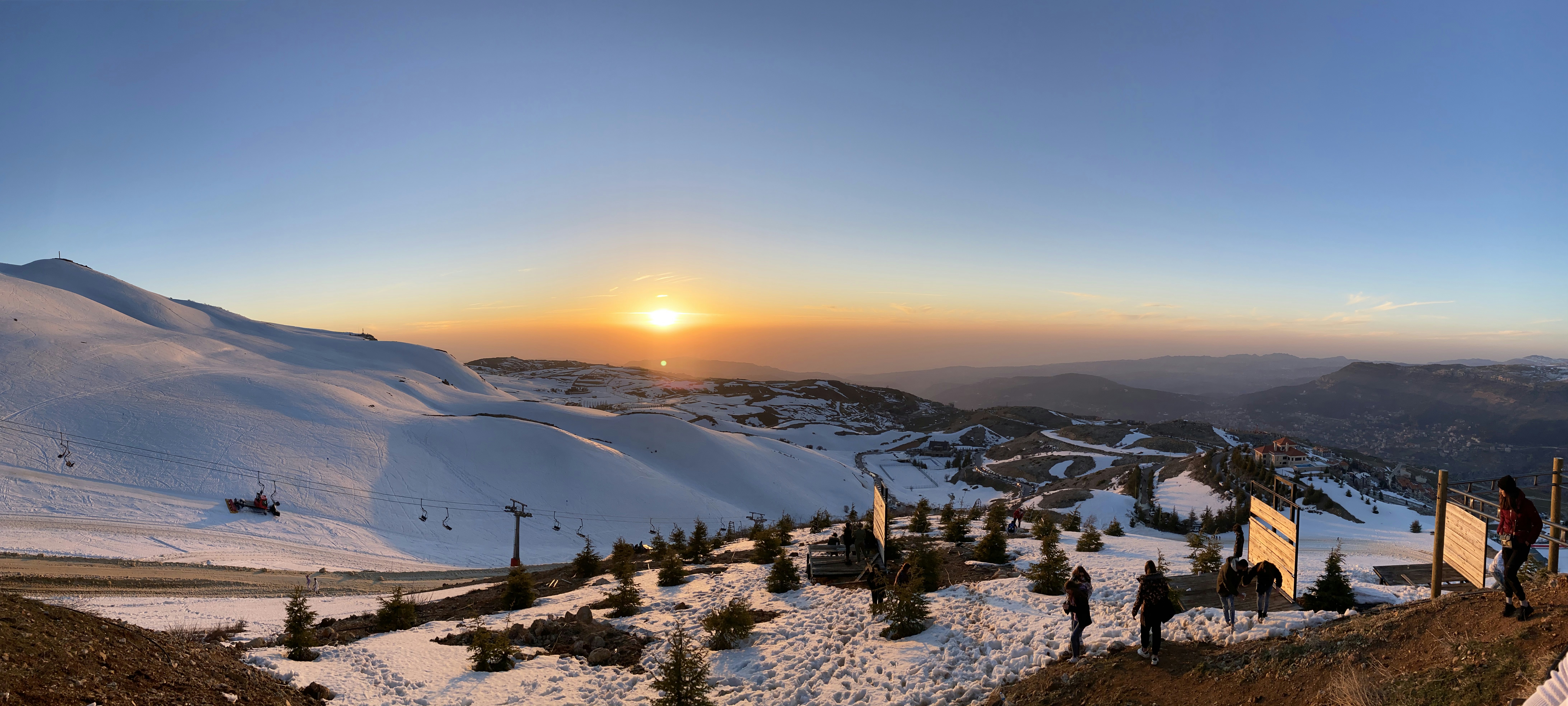 Golden sunset illuminating snow-covered hills with silhouettes of trees and people enjoying the serene landscape.