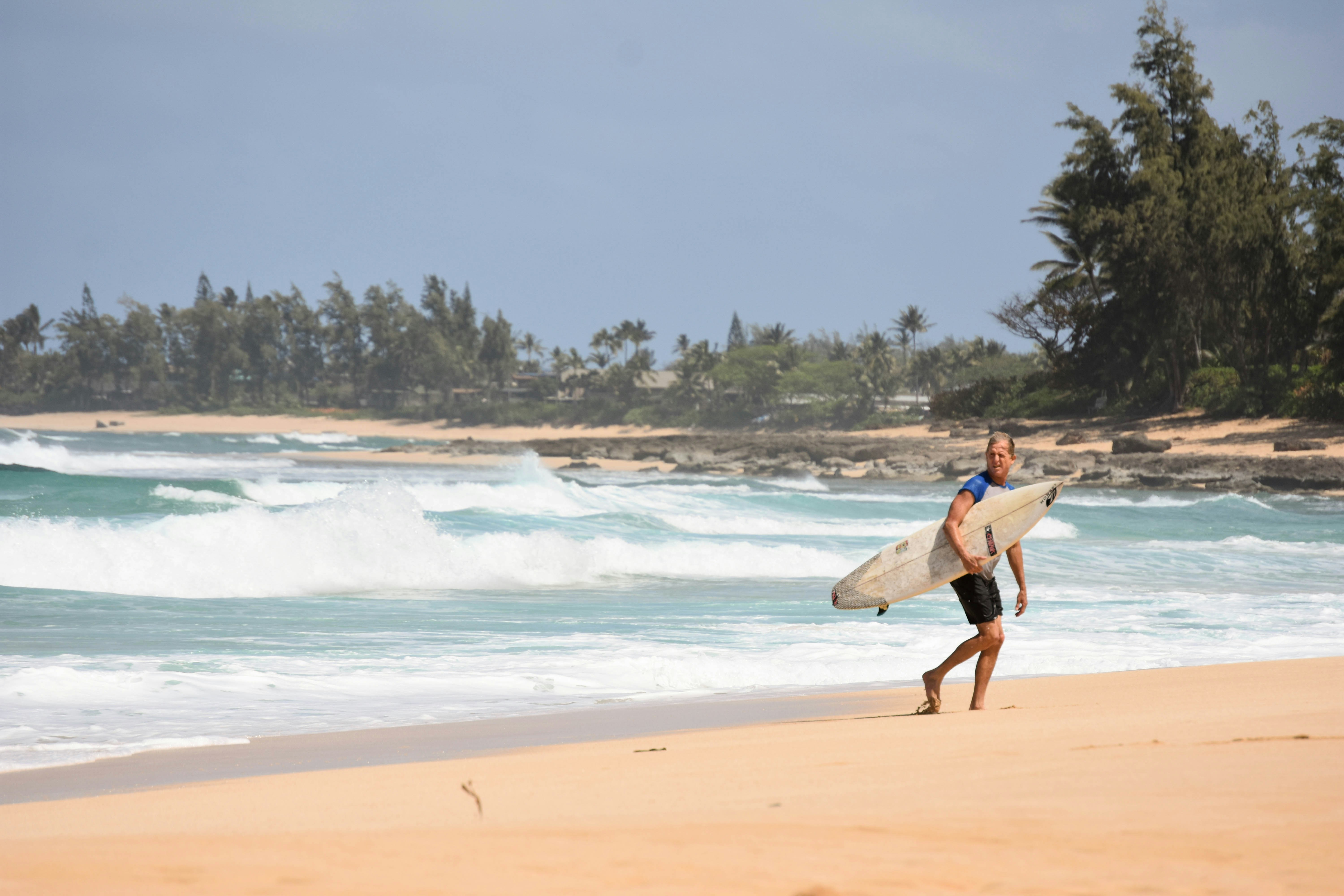 Shoreline Stride: A Surfer Returns to Sand Under Sunny Skies