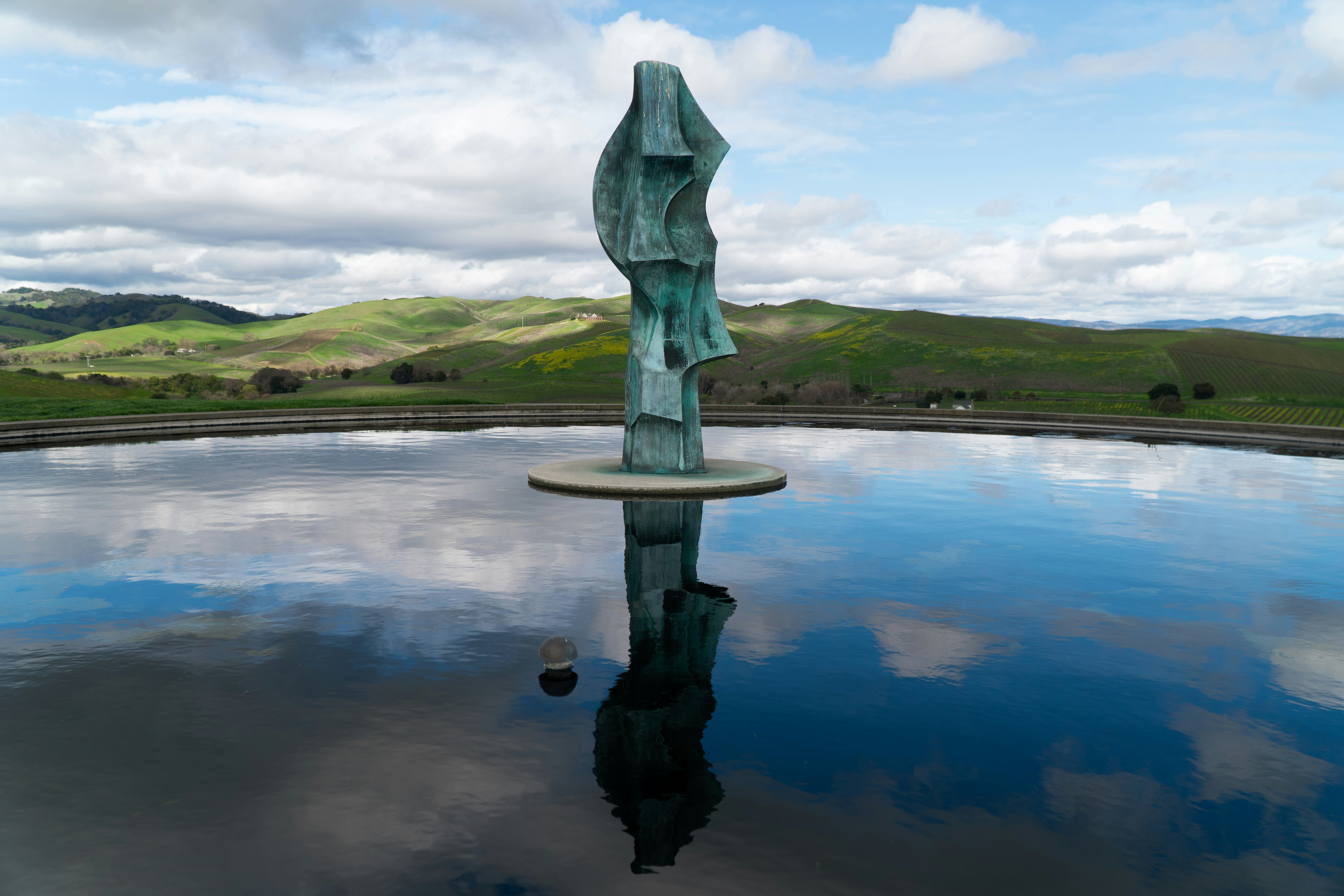 Abstract sculpture stands on a reflective water surface with green hills and cloudy sky in the background.