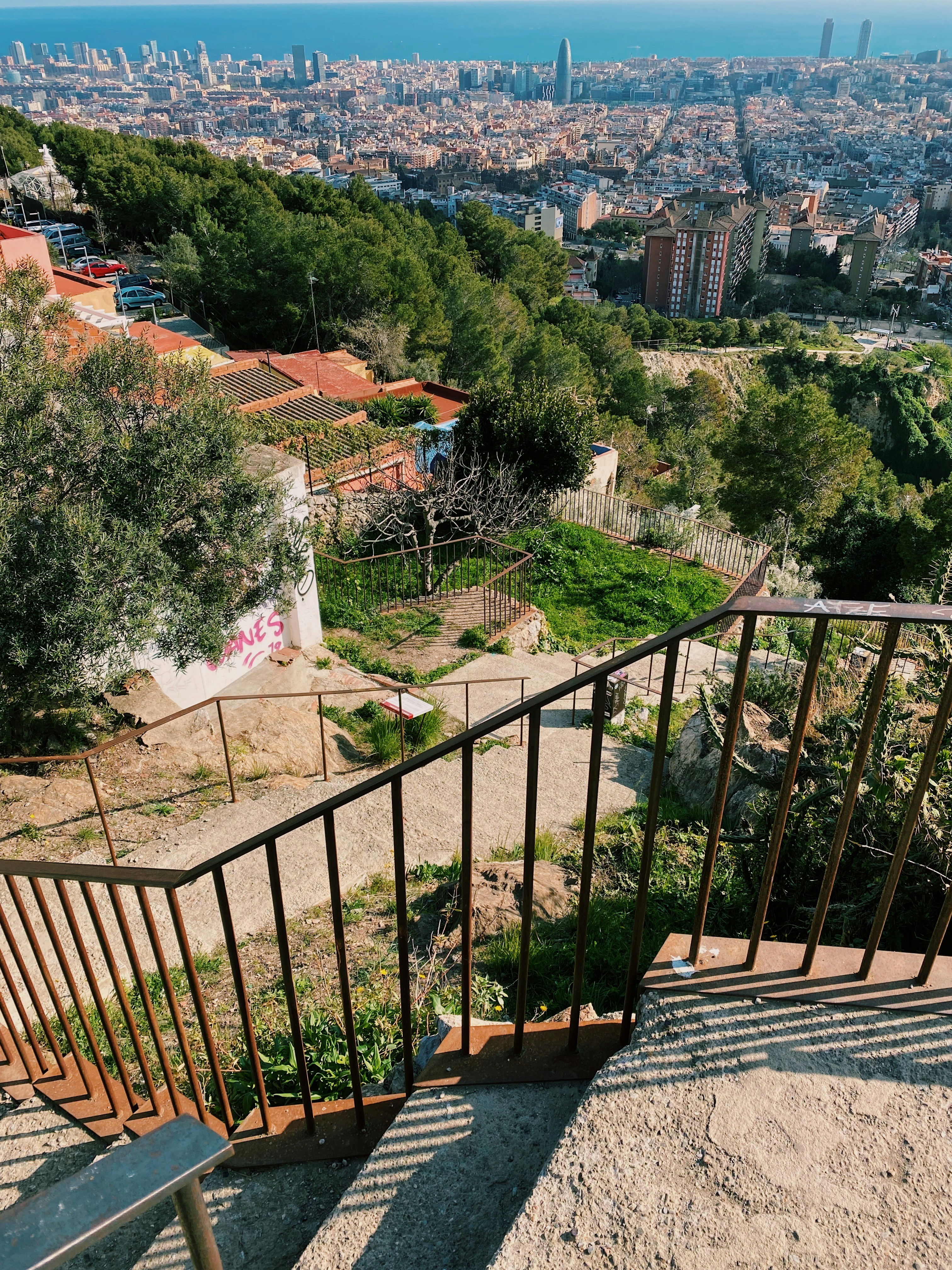 Winding pathway leading down a hillside, framed by greenery and overlooking a sprawling urban landscape with the sea in the distance.