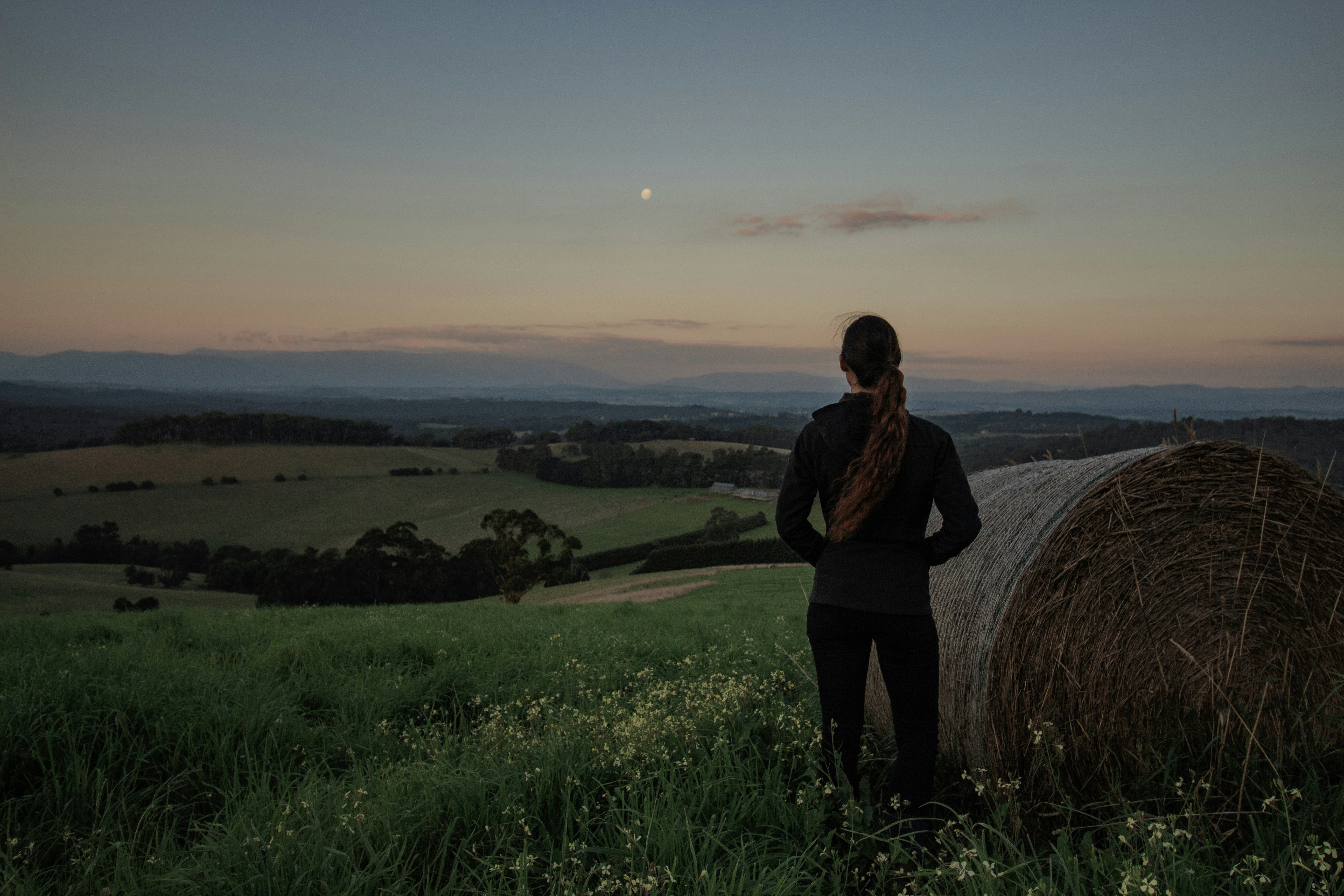 Silhouette of a woman in a black jacket gazing at a sunset over rolling green hills.