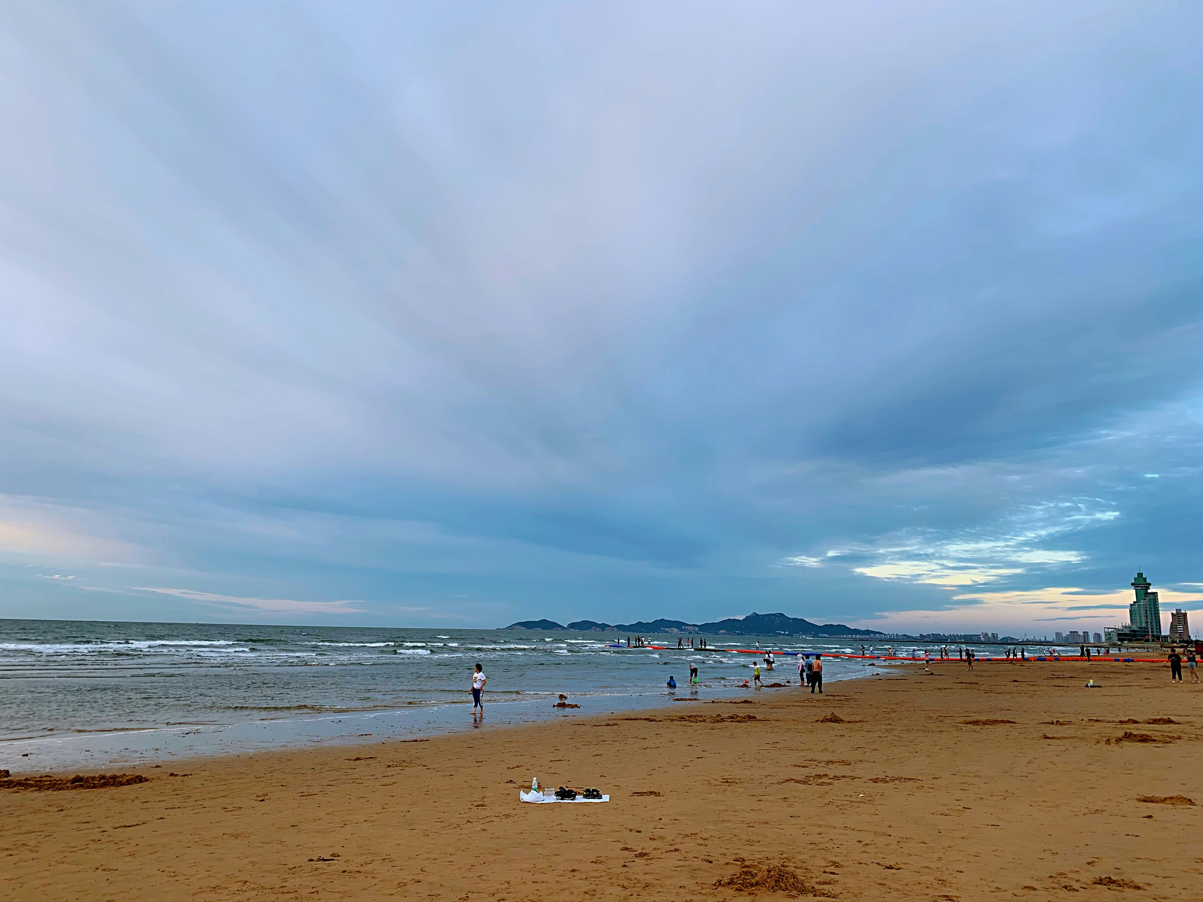 People strolling along a sandy beach at dusk, with gentle waves lapping at the shore and a distant lighthouse standing guard. 