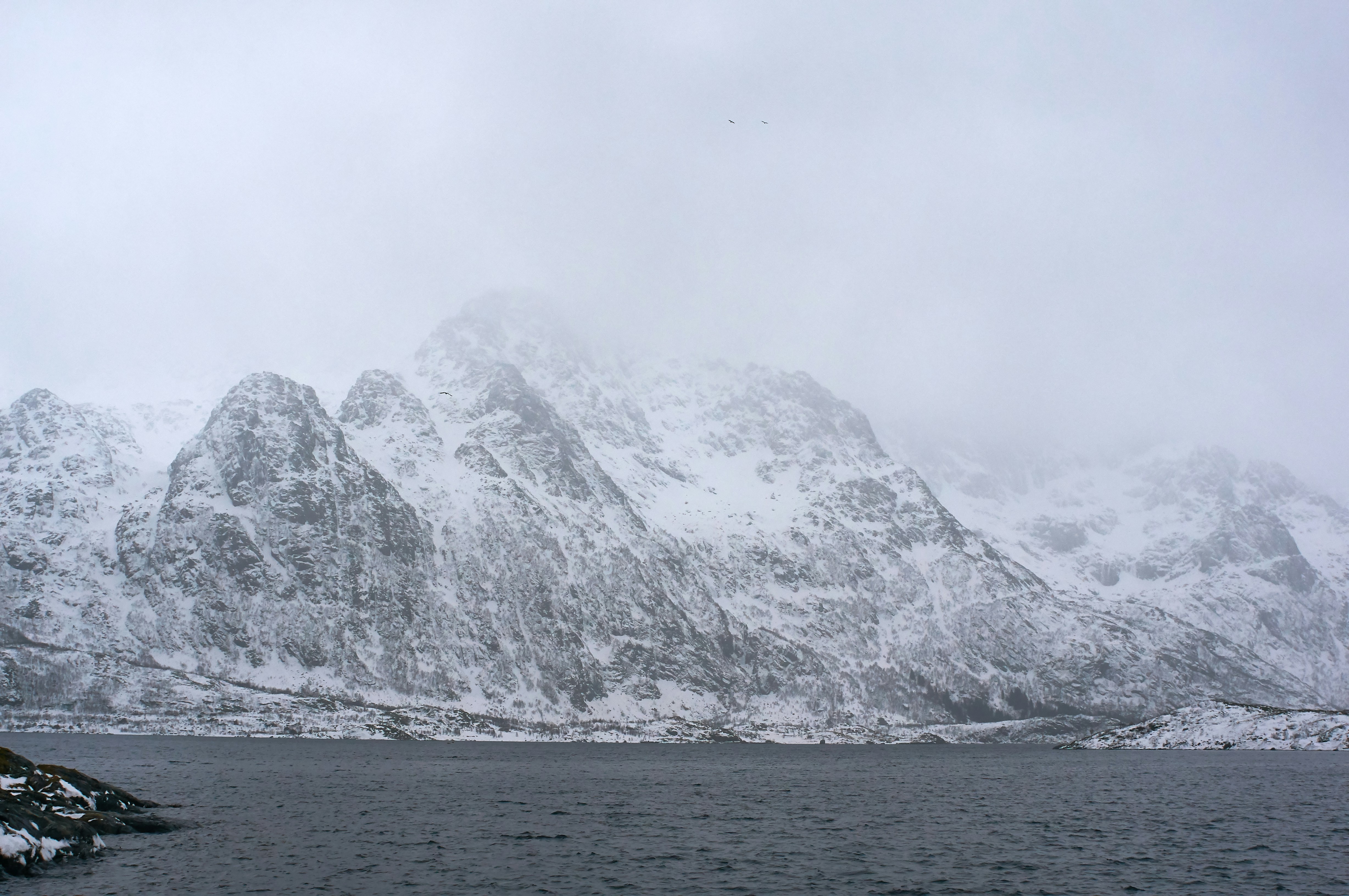 Snow-covered mountains shrouded in mist, partially reflected in a calm body of water. The scene evokes a sense of serene isolation.