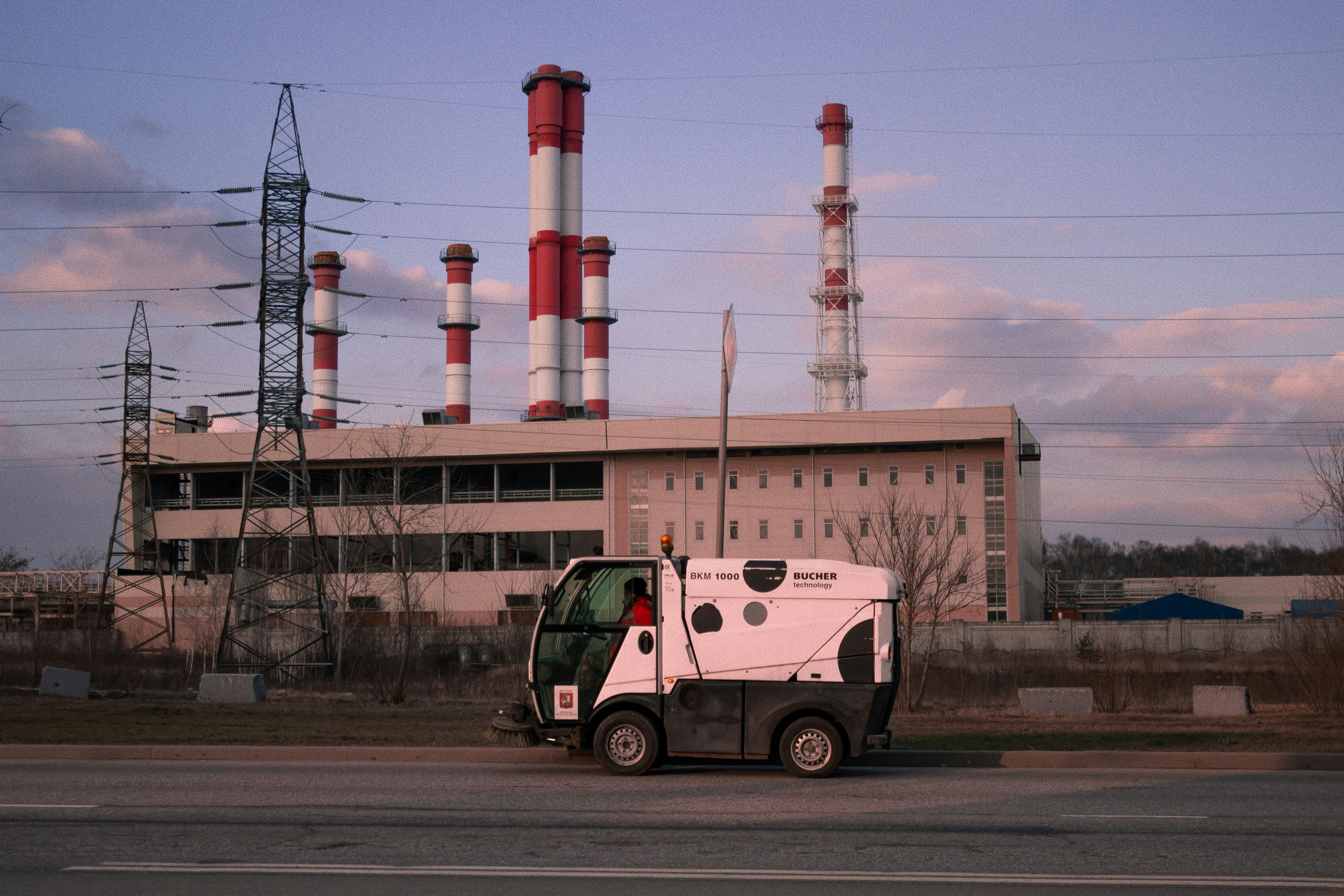 White street sweeper along a quiet road with red-and-white chimneys looming over the factory at dusk