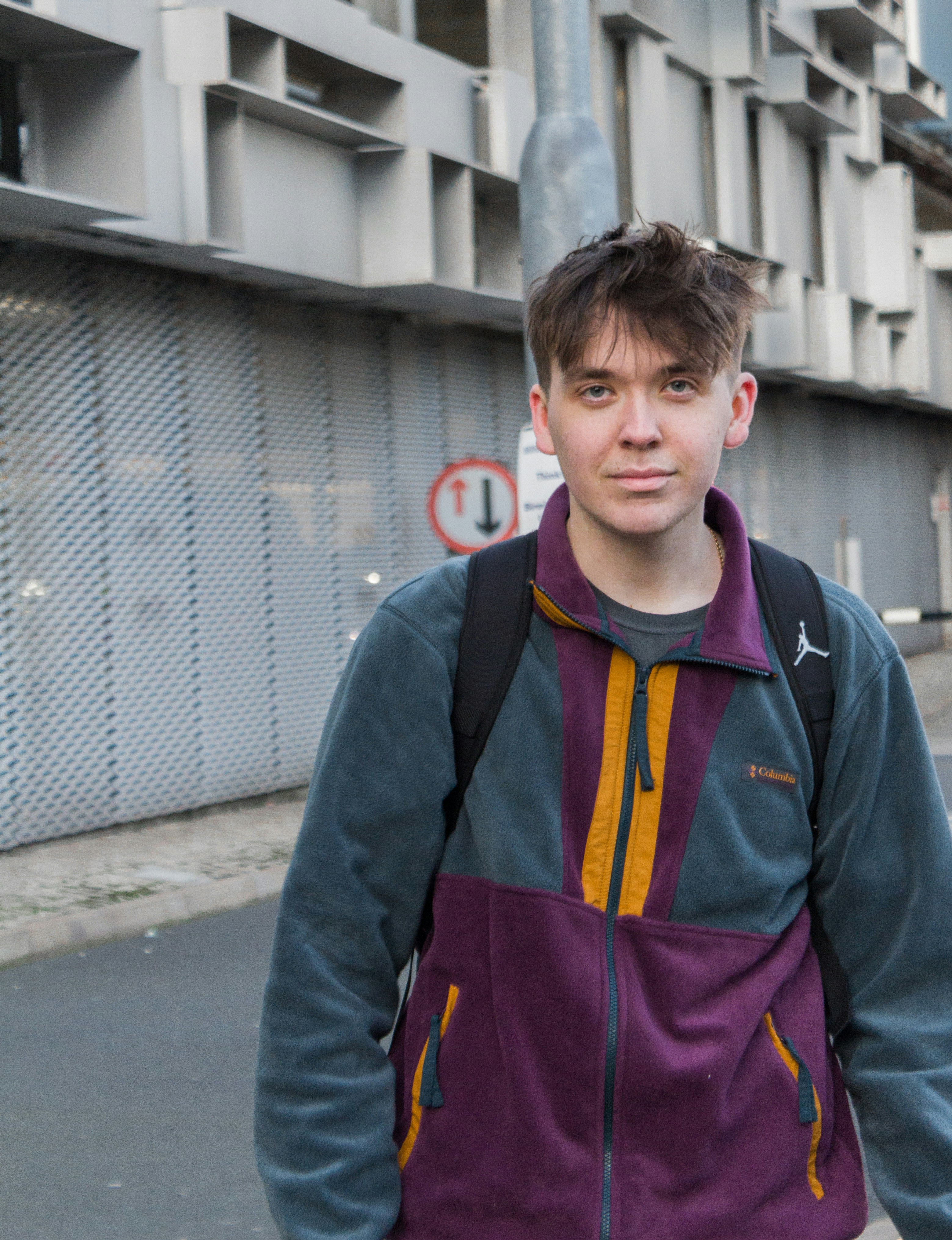 Young man in a colorful jacket standing on a city street, with a modern architectural backdrop and traffic signs visible.