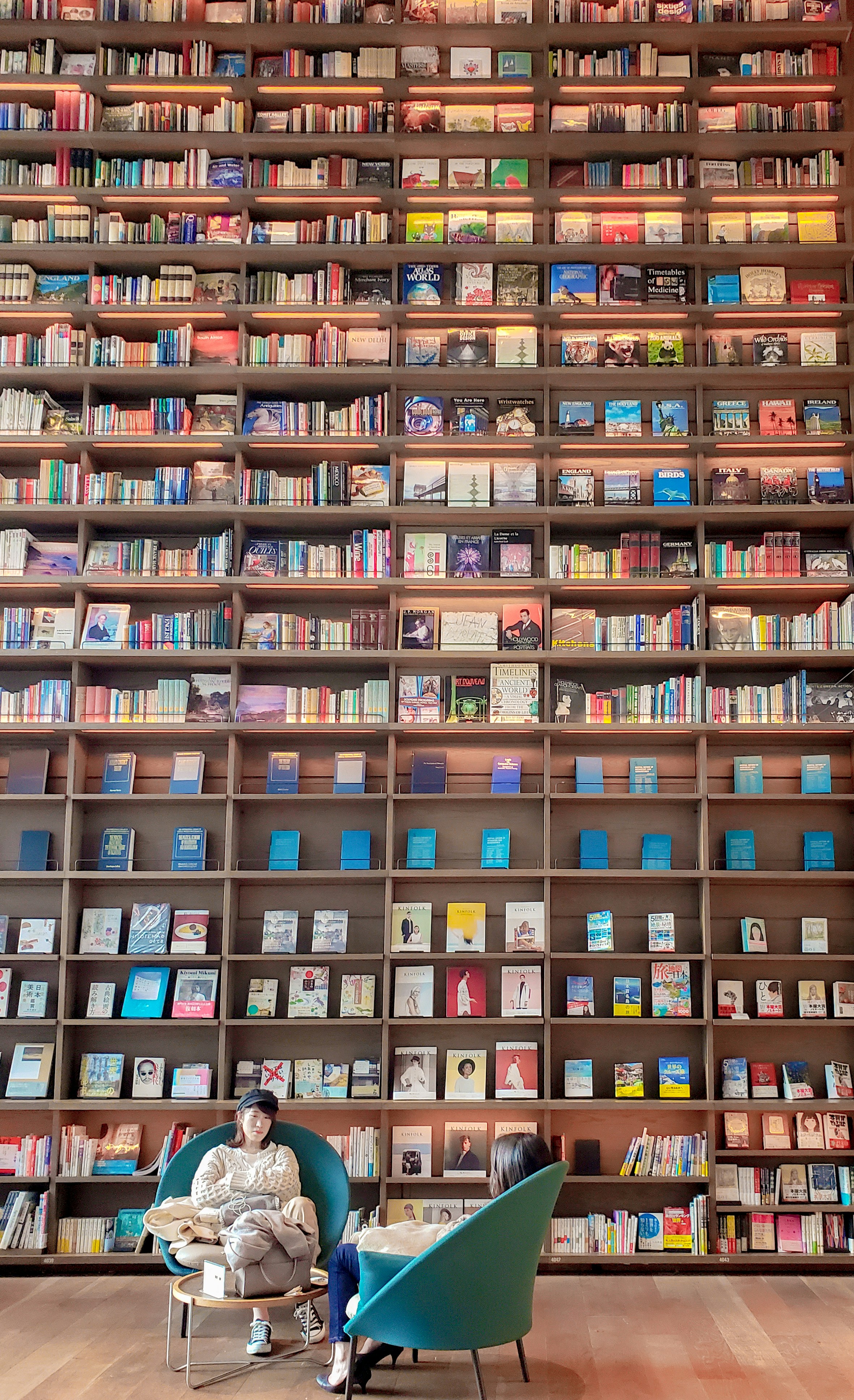 Two individuals relax in modern chairs in front of a towering bookshelf filled with colorful books. The inviting atmosphere encourages a love for reading.