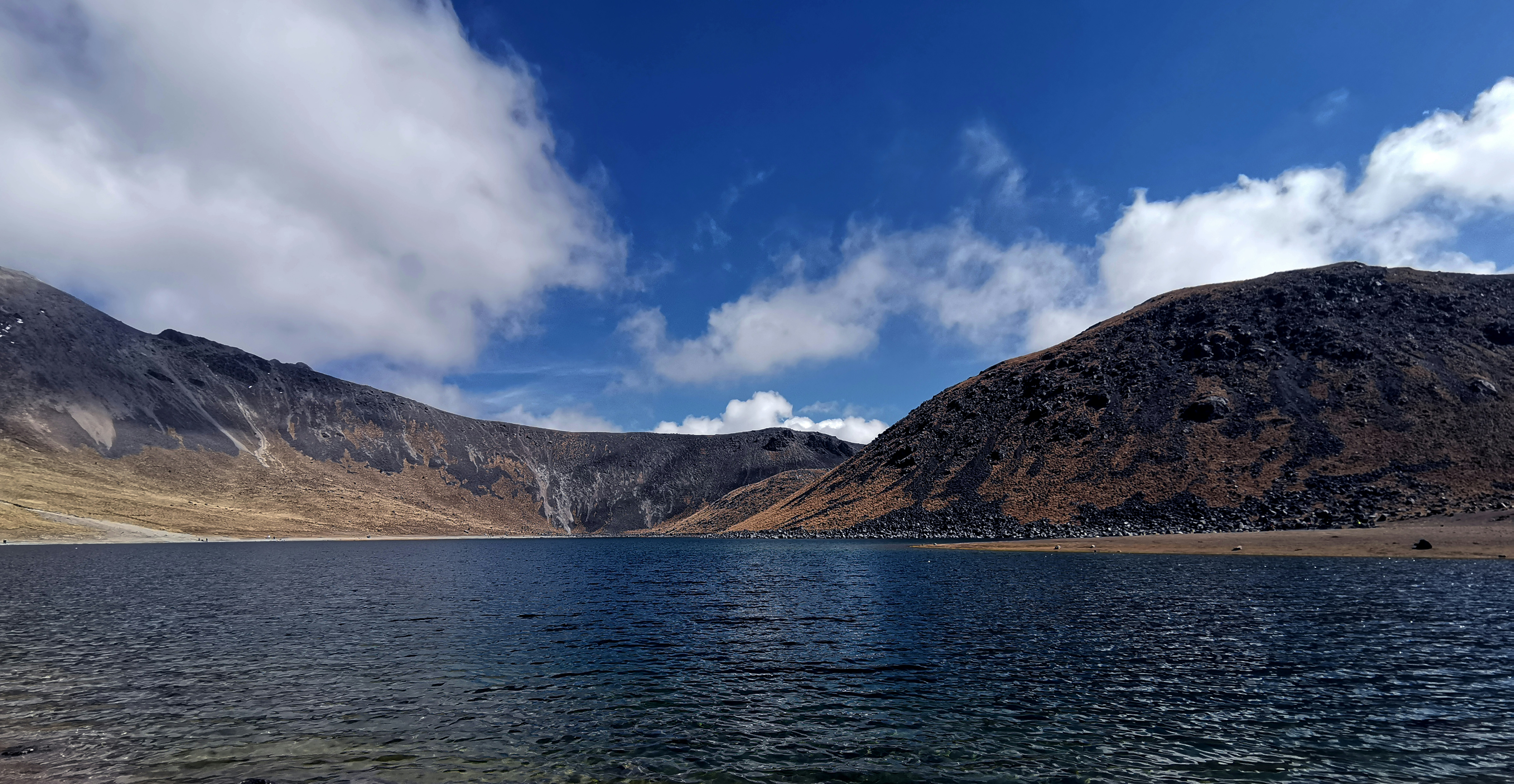 Clear mountain lake reflecting rugged peaks and a bright blue sky with scattered clouds.