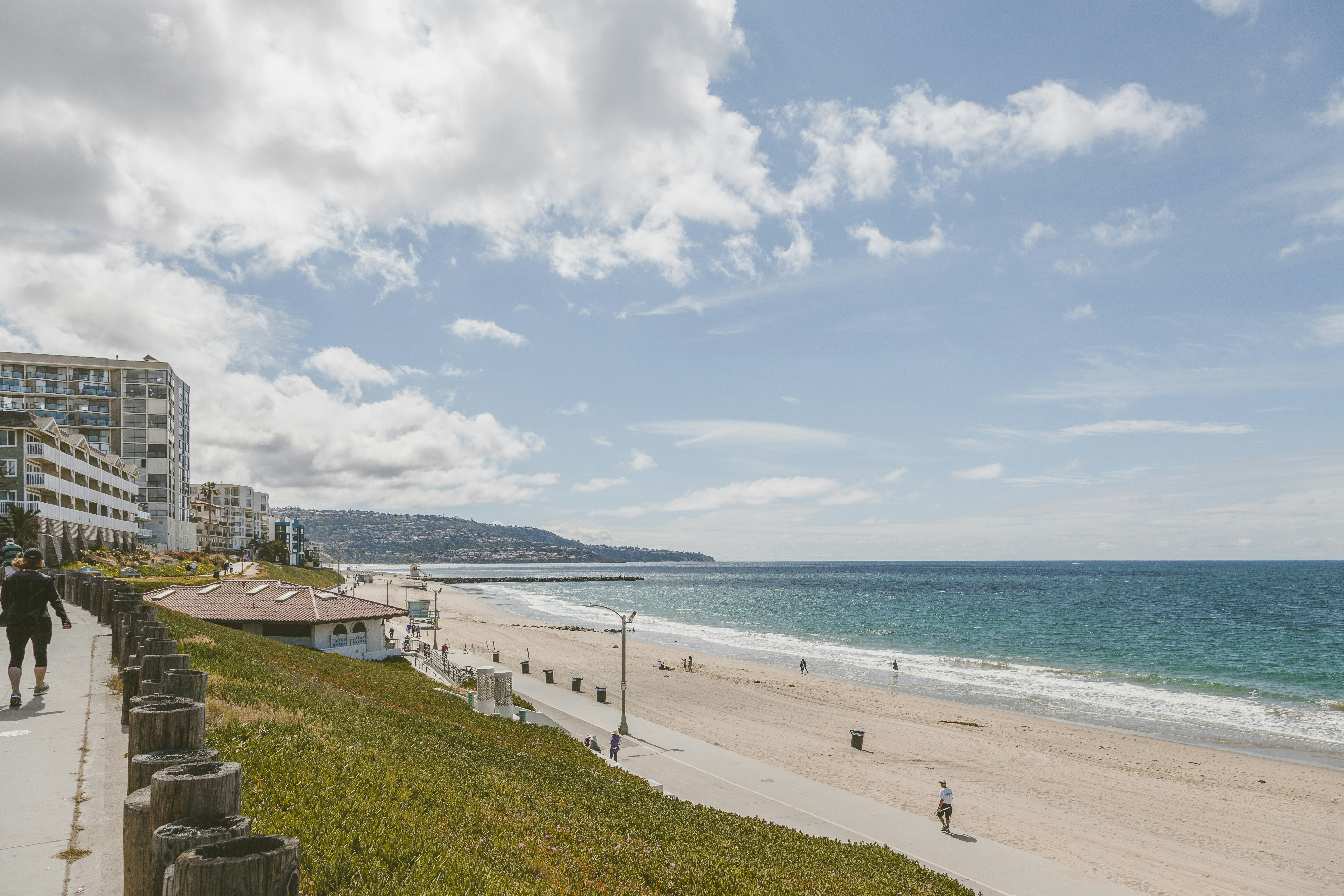 Sunny beach view with ocean waves, coastal buildings, and hills in the distance.