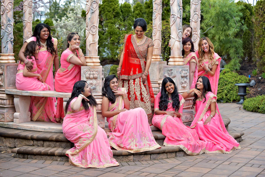 Group of women in bright pink sarees gathered around an ornate stone pavilion in a garden. One woman in a red-and-gold outfit stands at the center while the others pose seated and smiling.