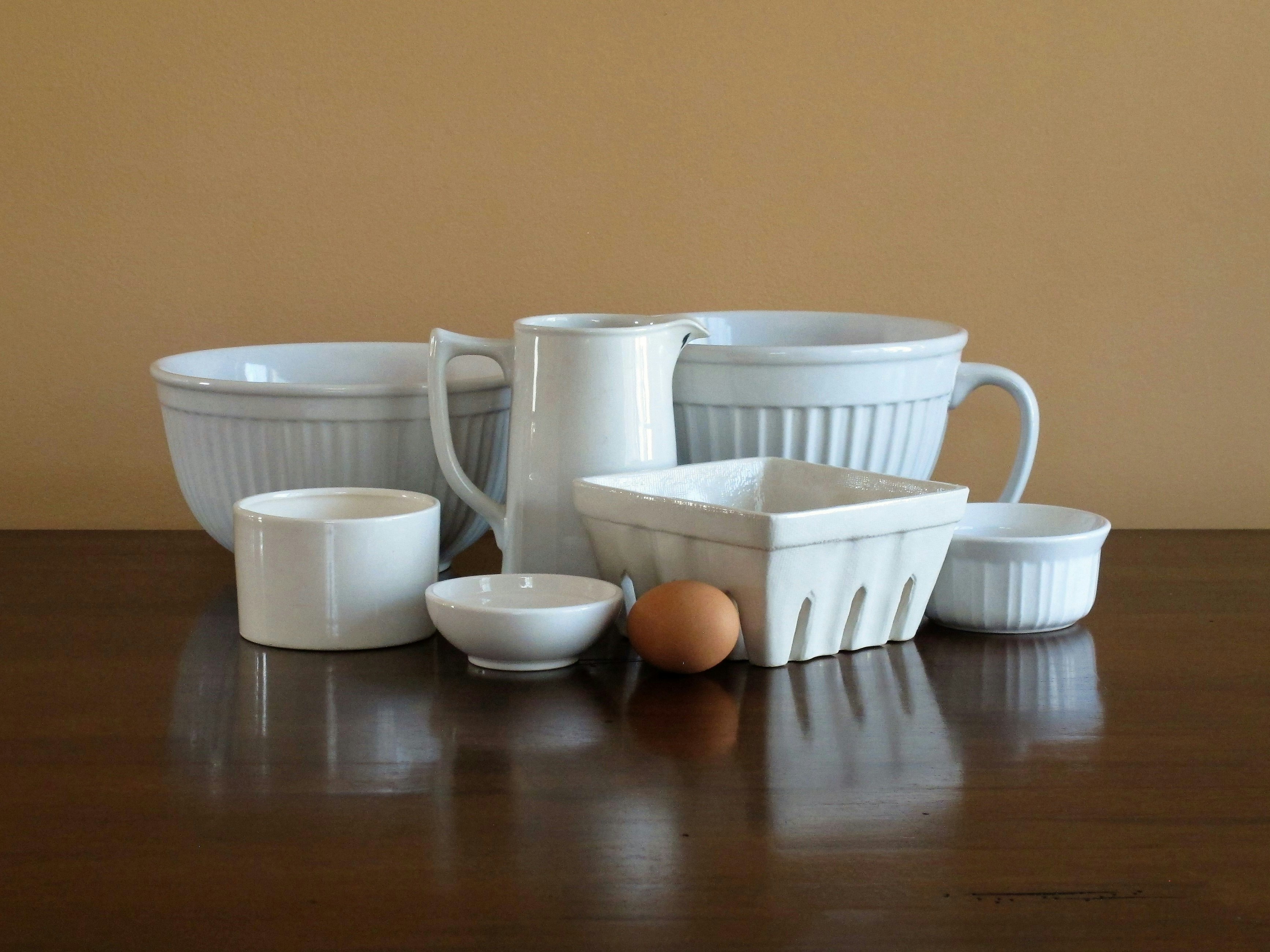 White ceramic bowls, a pitcher, and an egg arranged on a brown wooden table.