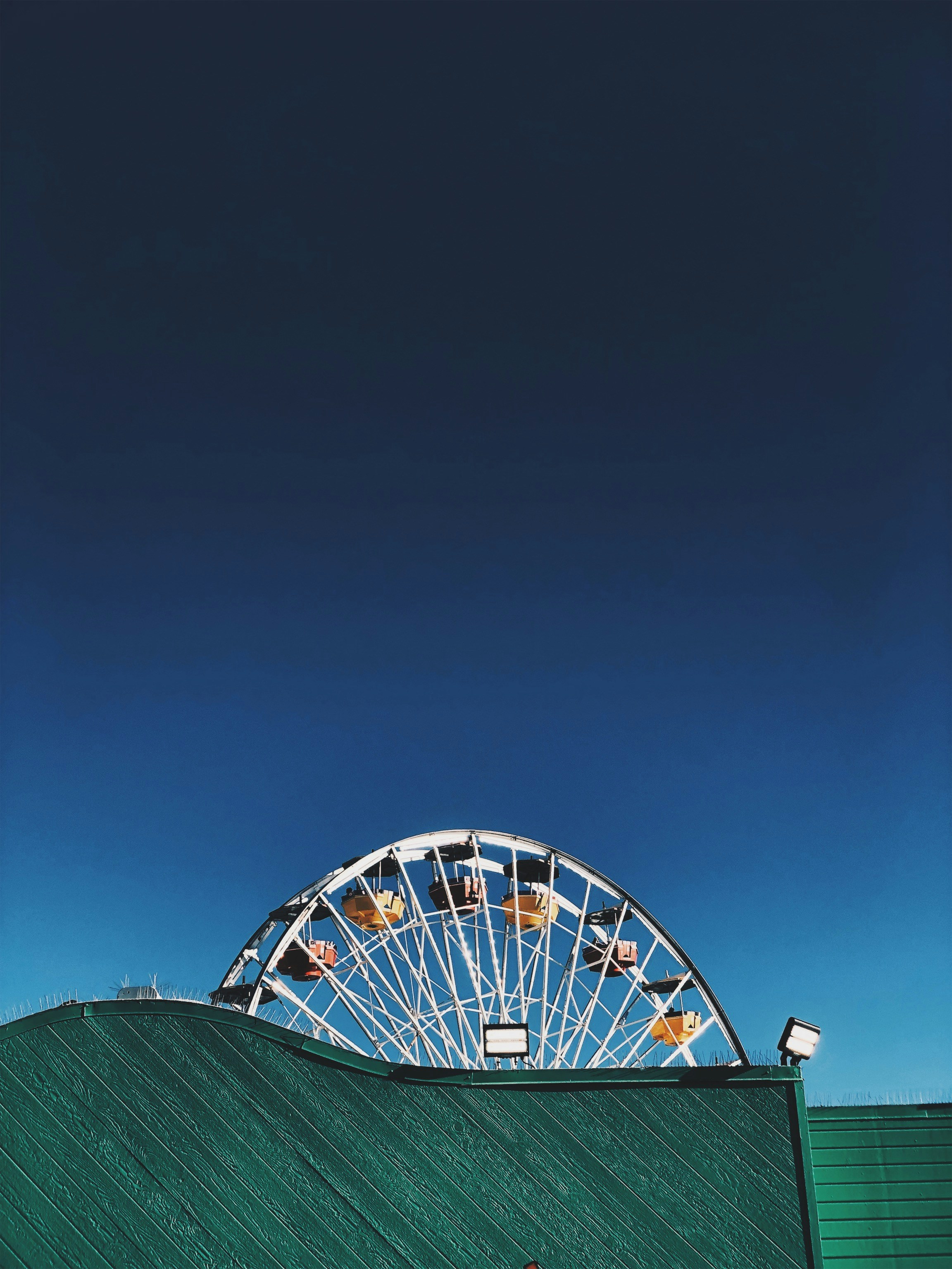 A vibrant Ferris wheel stands against a clear blue sky, framed by a textured green structure below. The scene evokes a sense of nostalgia and excitement.
