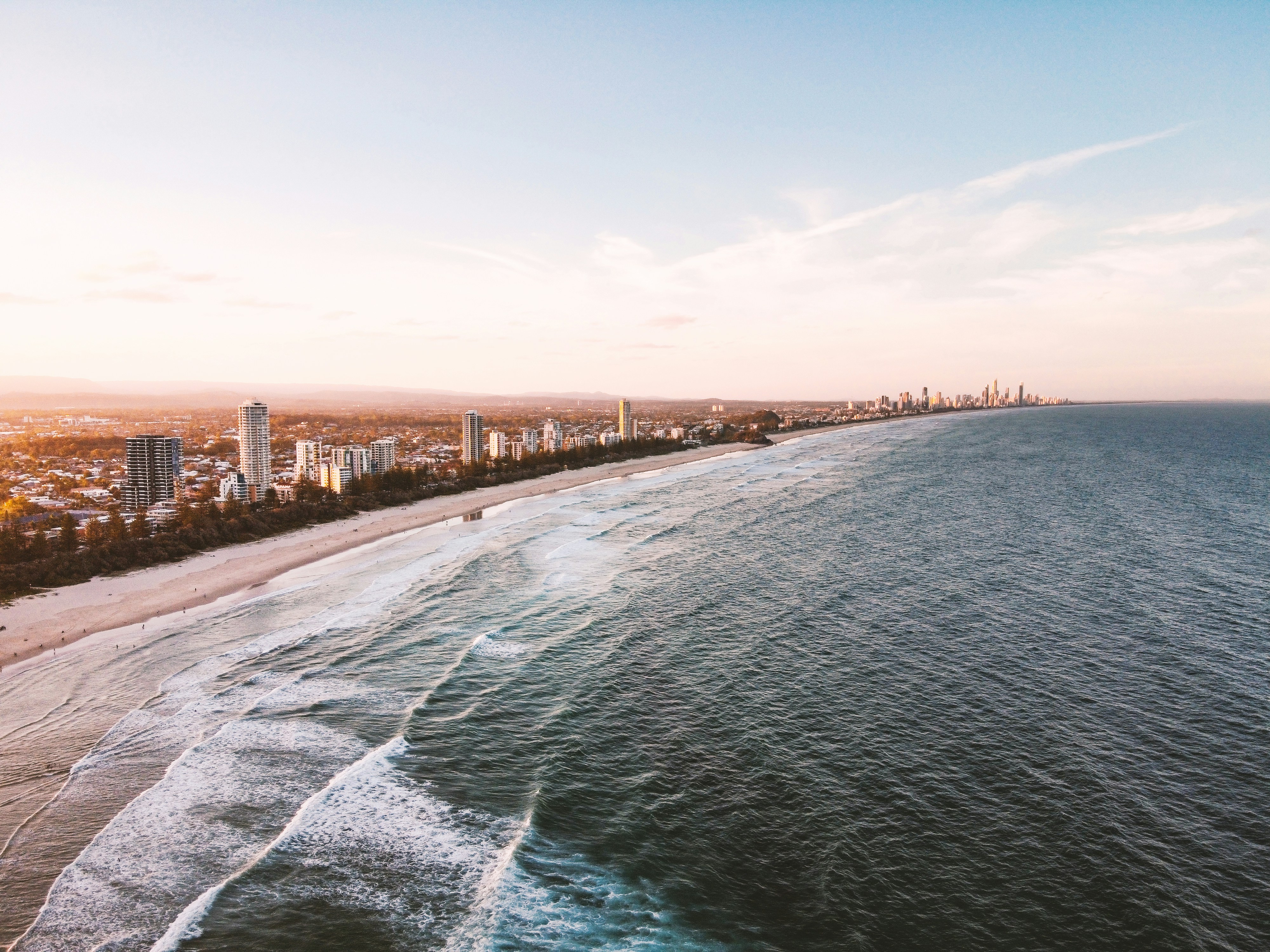 Aerial view of a tranquil beach with gentle waves lapping against the shore, framed by a city skyline in the distance during sunset.
