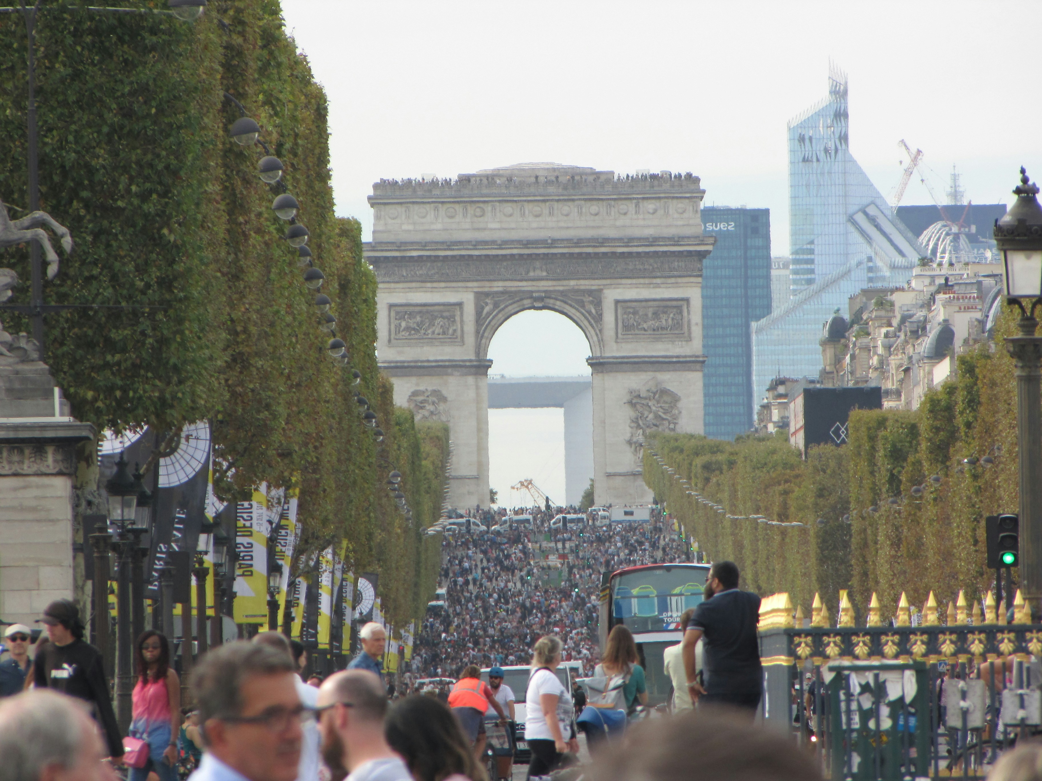 Crowded Champs-Élysées spill toward the Arc de Triomphe, captured in a daytime photograph with trees lining the avenue and a distant bus.