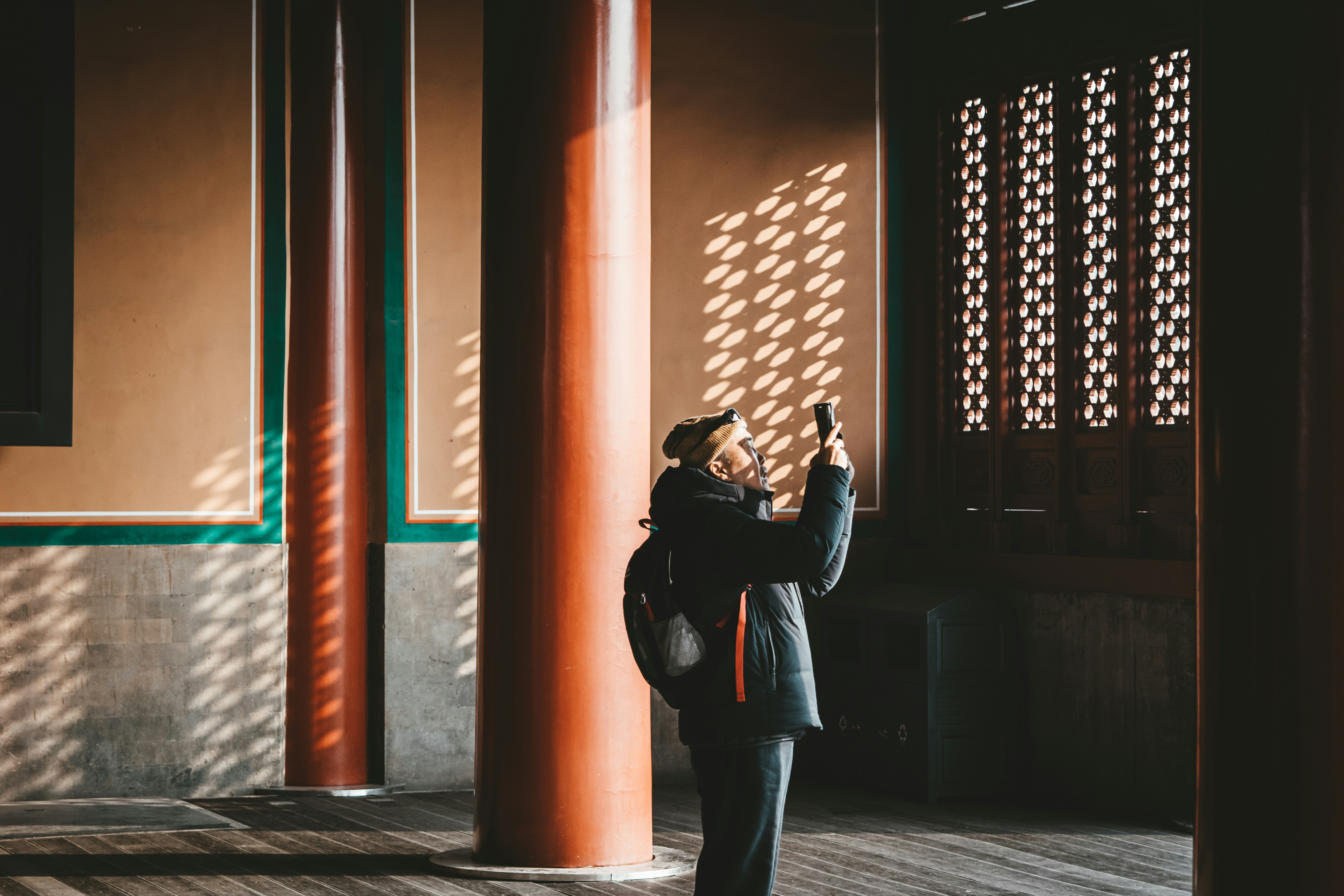 Person photographing intricate shadows cast by ornate window lattice in a sunlit hallway.