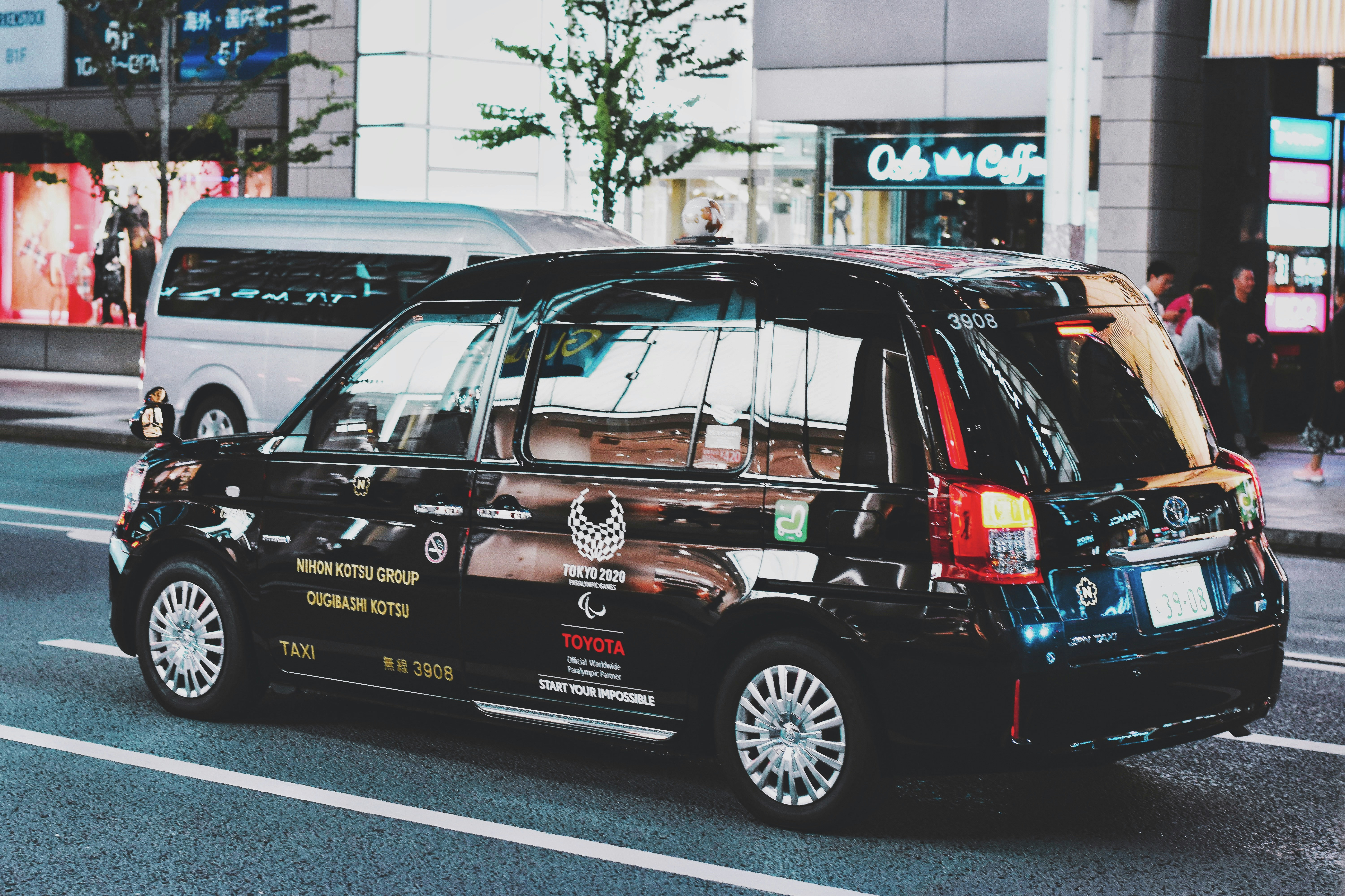 Black taxi with Olympic branding navigating a busy Tokyo street, showcasing urban life and transportation. The scene captures the essence of city dynamics.