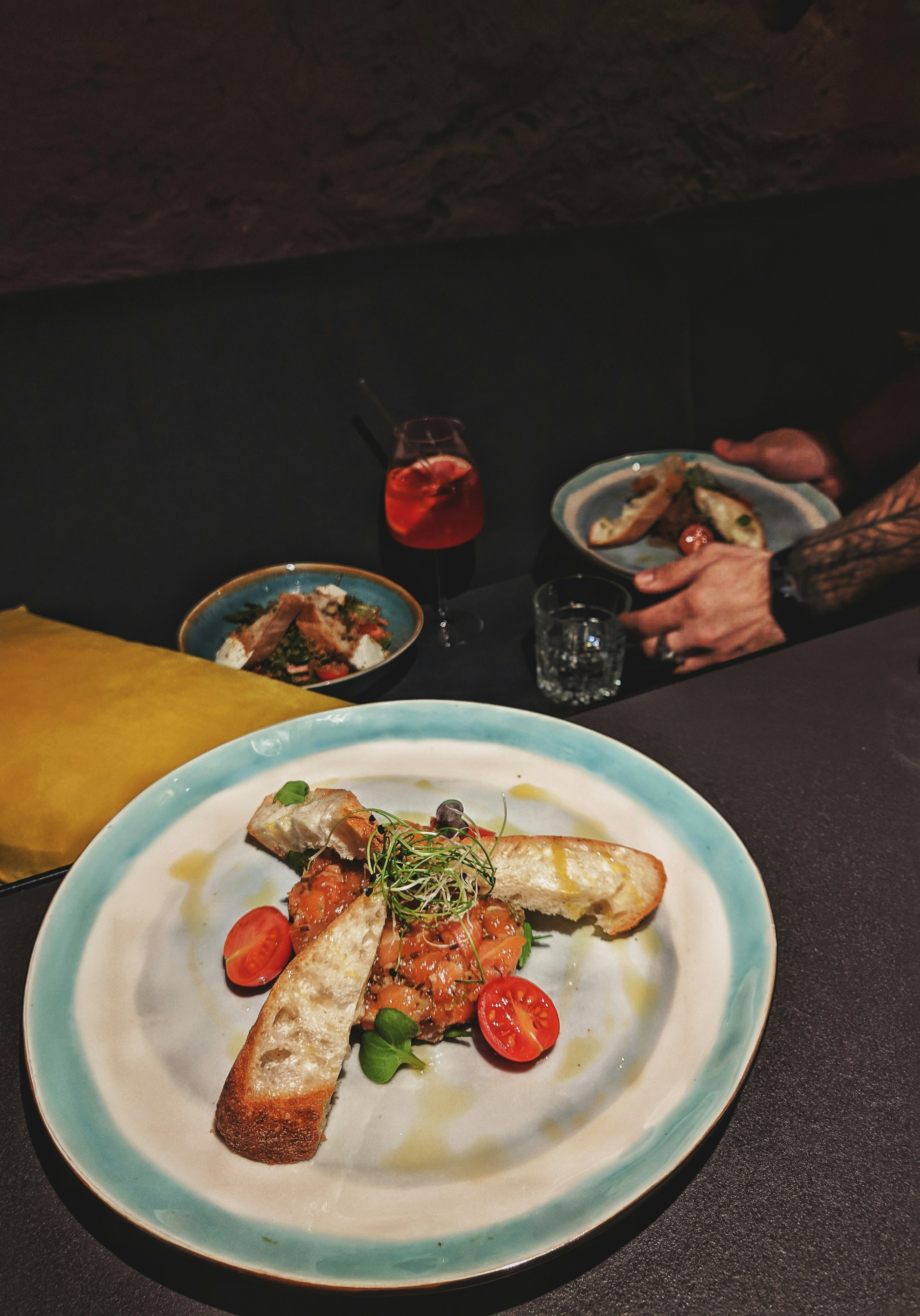 Artfully arranged dish featuring fresh ingredients, accompanied by a vibrant cocktail and a hint of a second plate in the background.