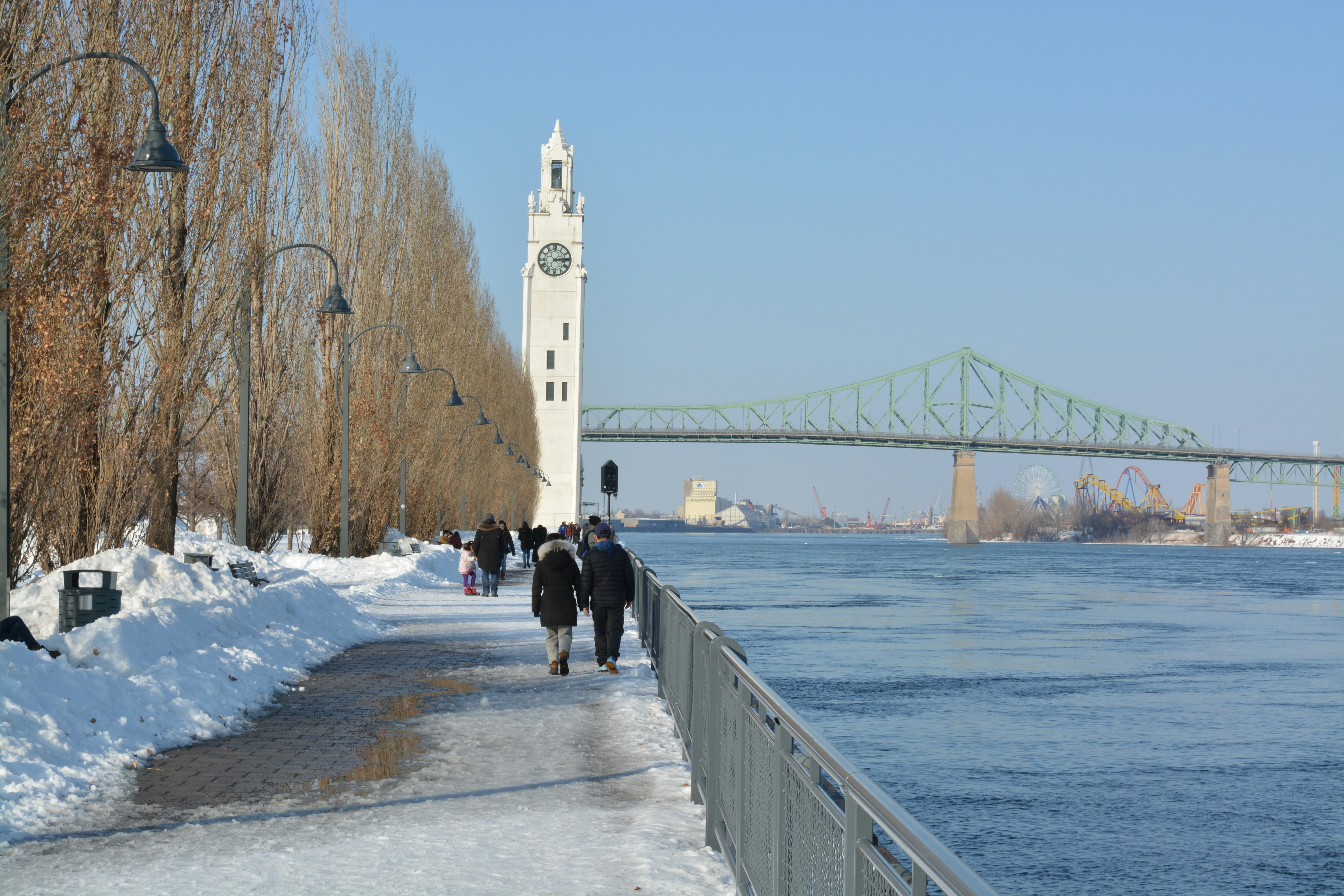 Snow-covered pathway lined with trees and a historic clock tower, with a bridge spanning the river in the background.
