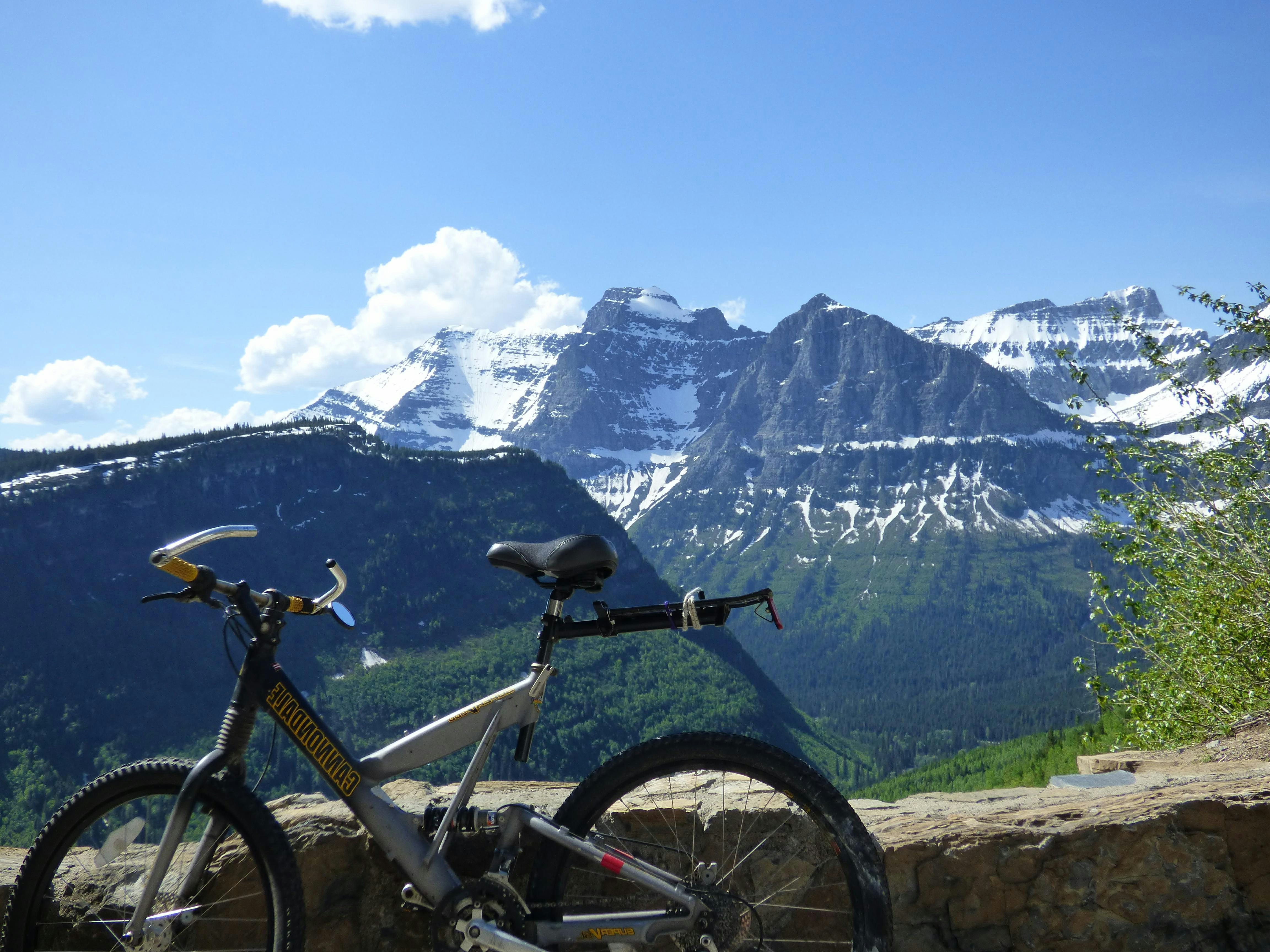 Mountain bike resting on a rocky ledge with snow-capped peaks and vibrant greenery in the background.