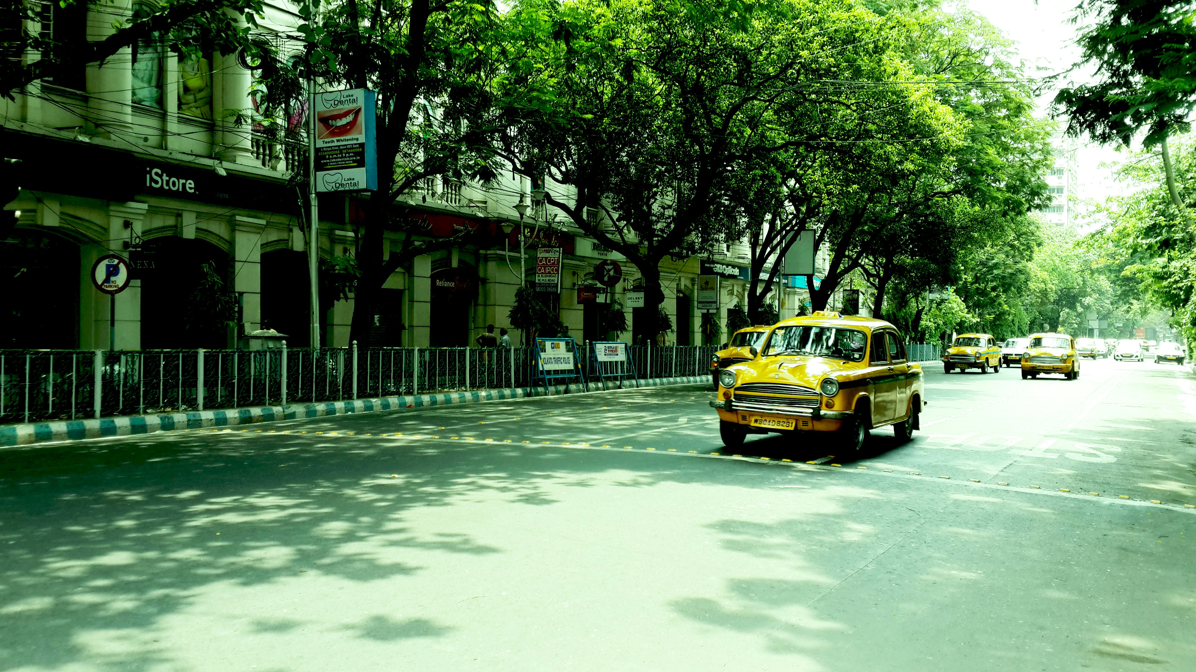 Classic yellow taxi navigating a sunlit street lined with lush trees and urban storefronts.