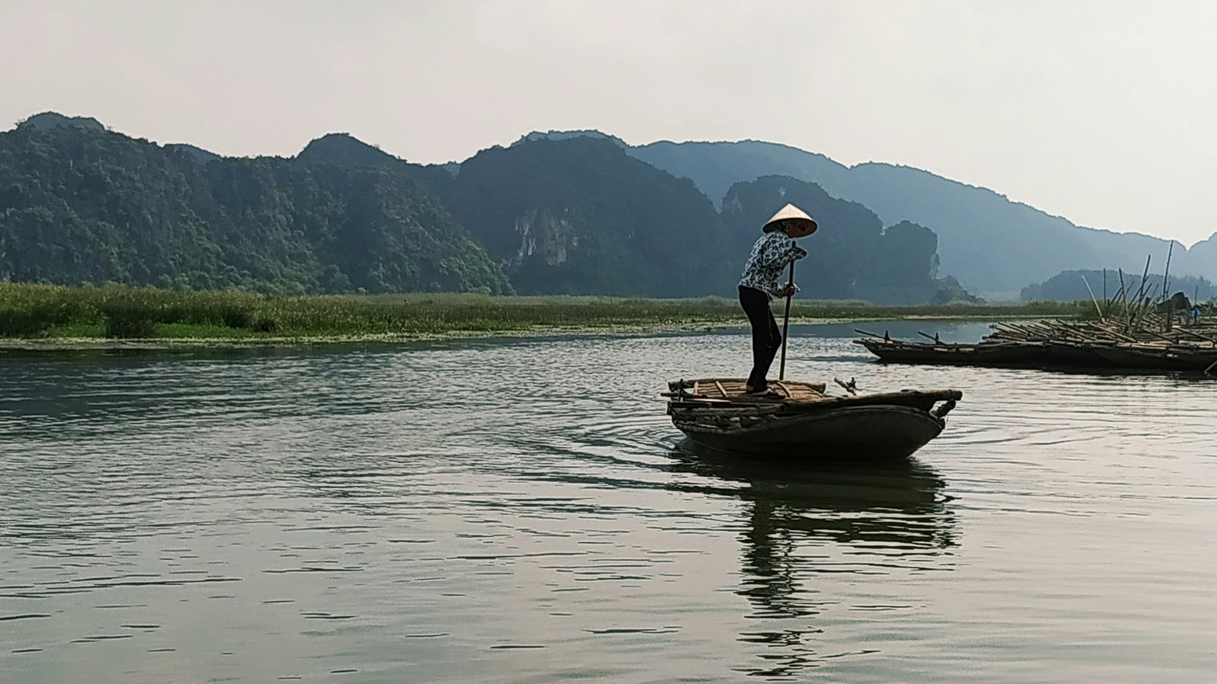 Fisherman standing on a traditional wooden boat, skillfully paddling through calm waters surrounded by lush mountains. 
