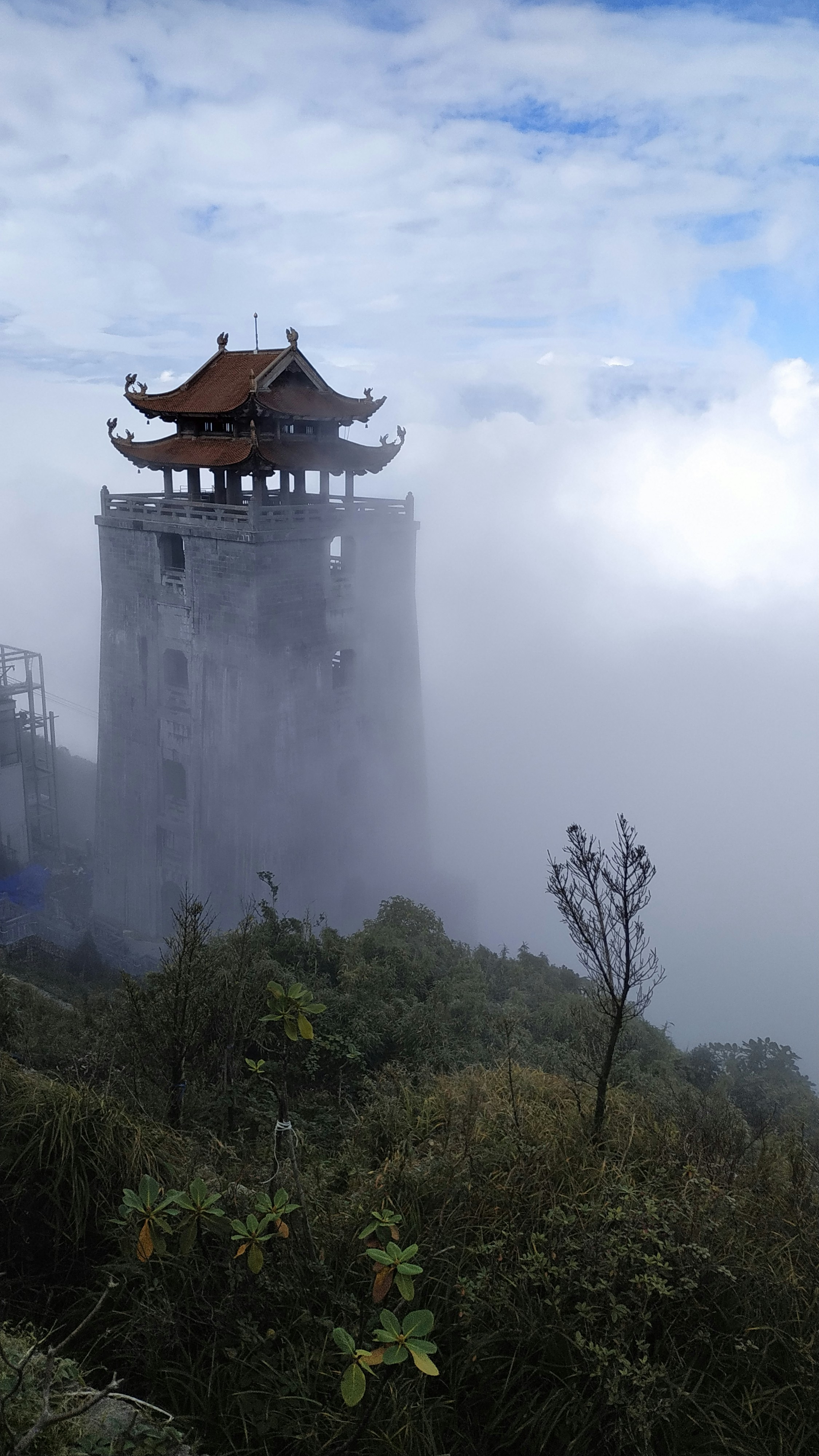 Ancient tower emerging from thick fog atop a mountain, surrounded by lush greenery.
