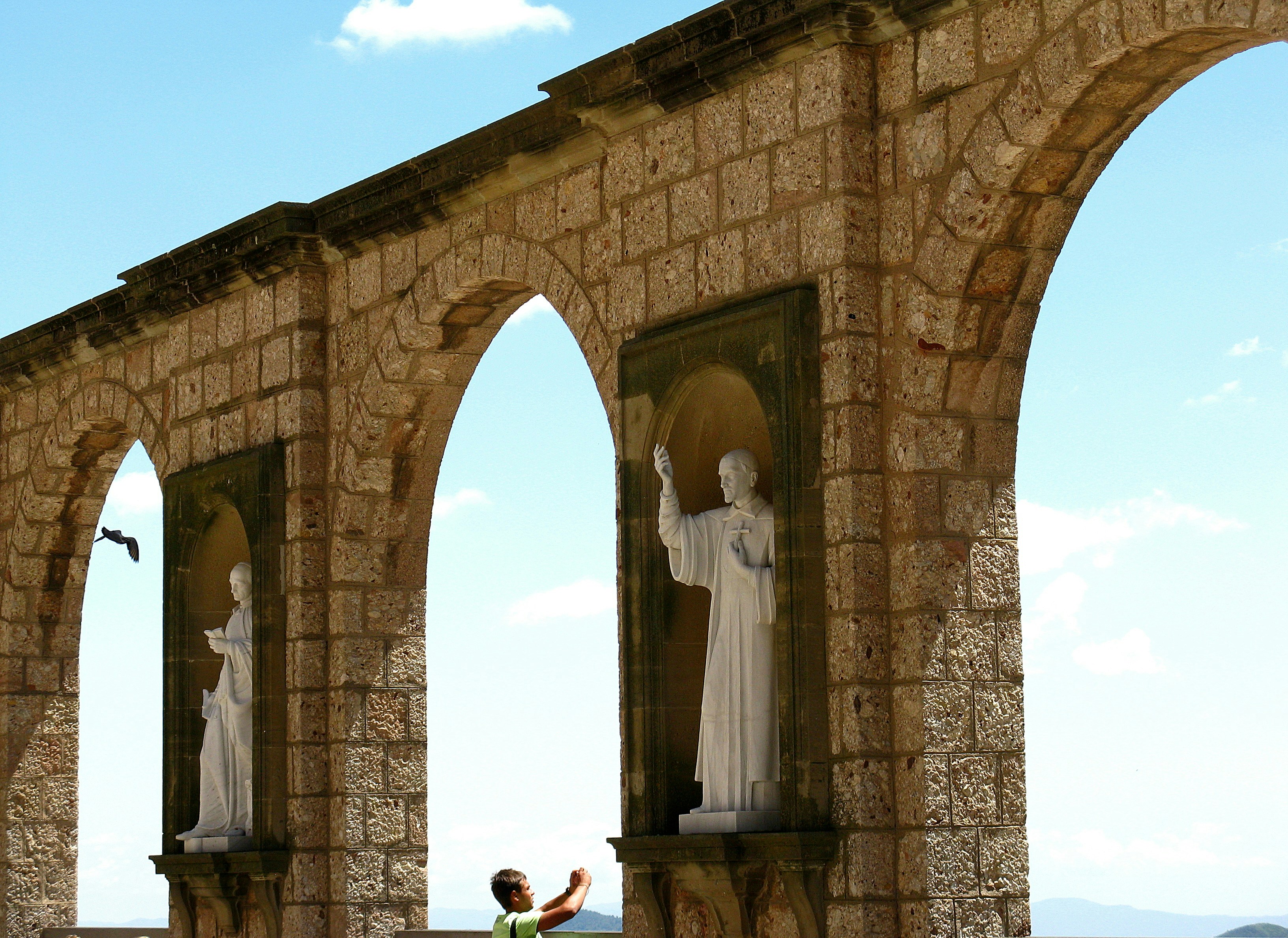 Weathered stone arches frame statues in alcoves along a seaside arcade, with a person at the base near the central niche under a bright blue sky. Photograph.