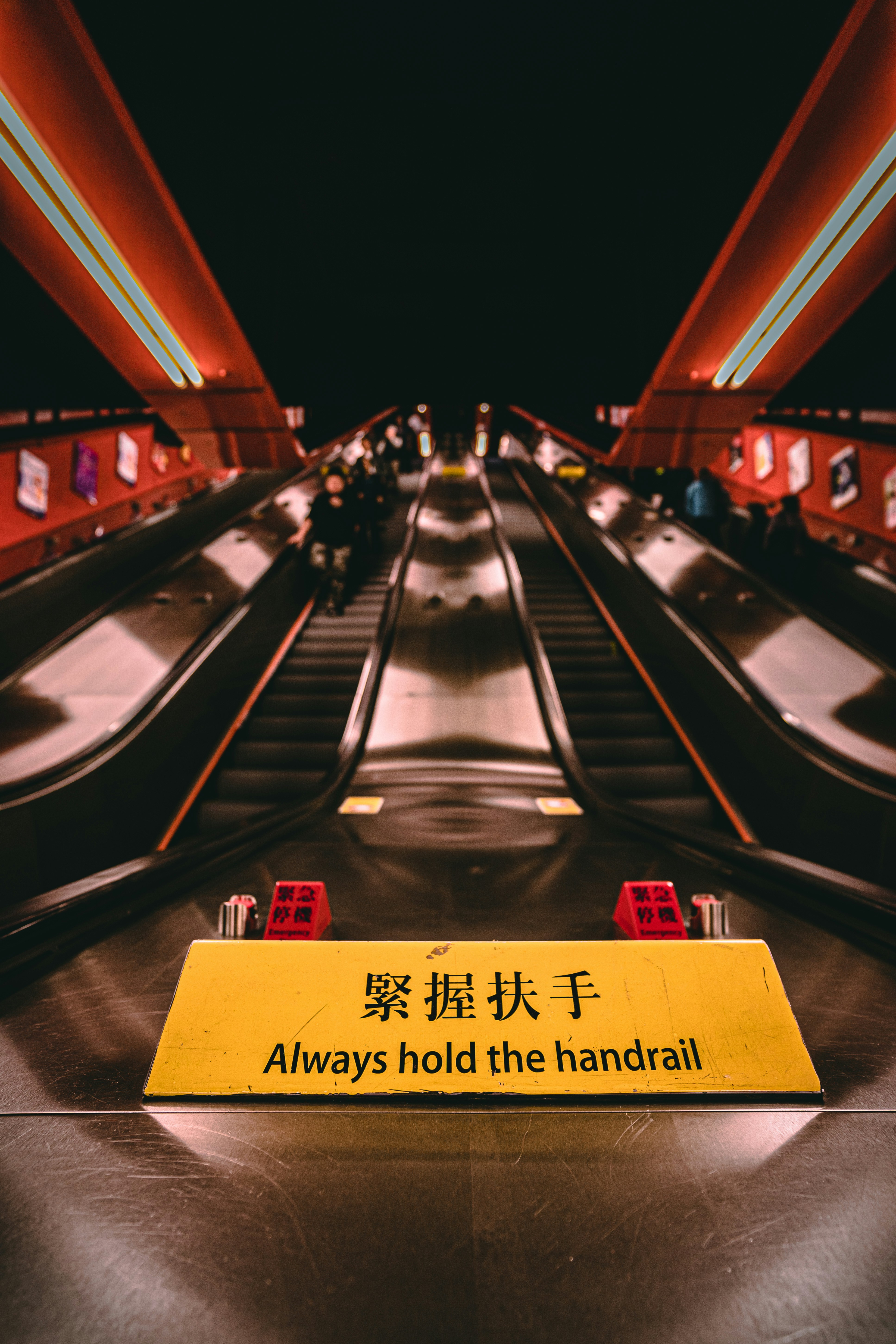 A yellow caution sign instructs passengers to hold the handrail on an escalator, framed by the vibrant architecture of the station.