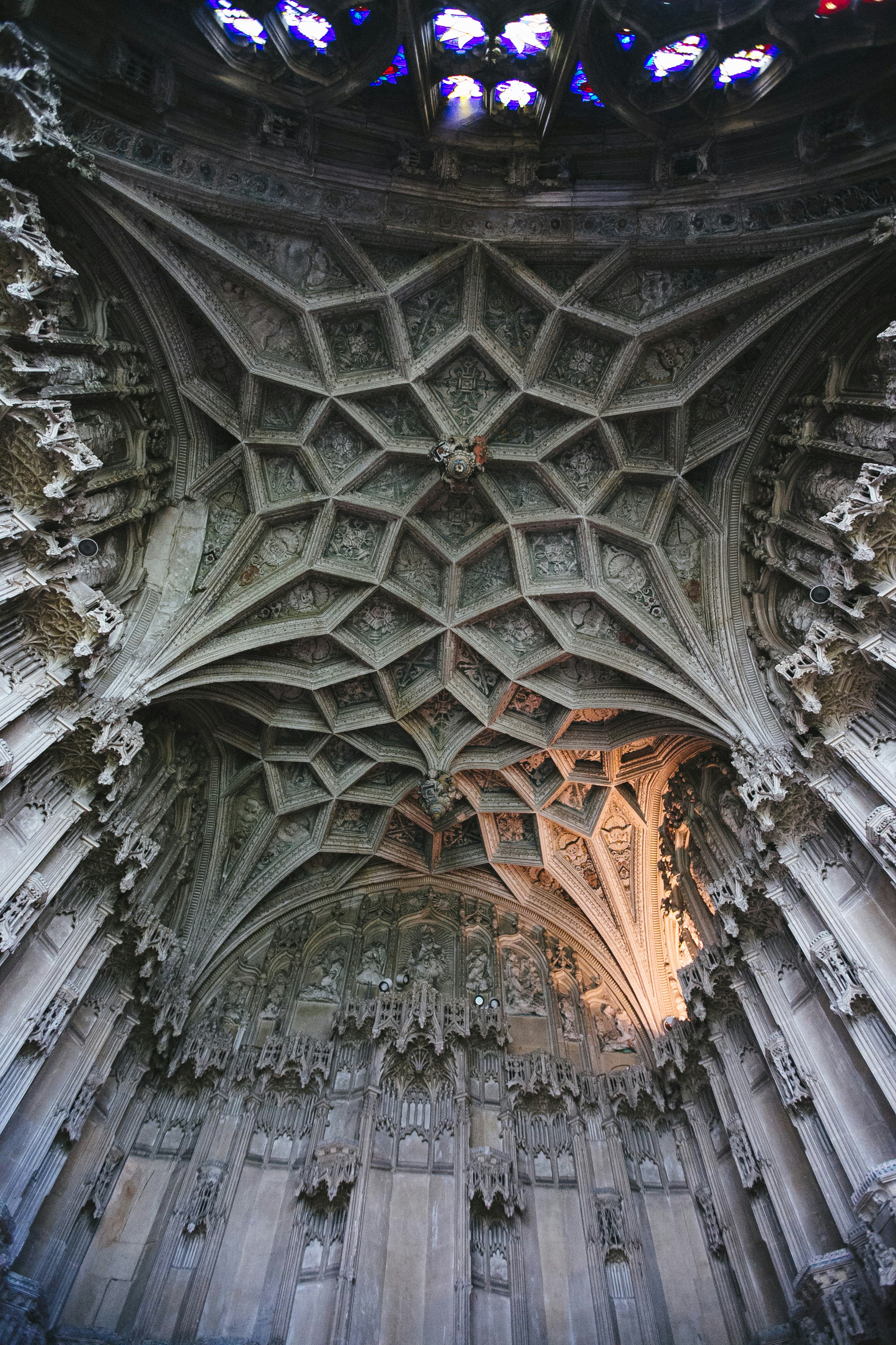 Intricate Gothic ceiling featuring elaborate stone carvings and stained glass elements. The design creates a mesmerizing interplay of light and shadow.
