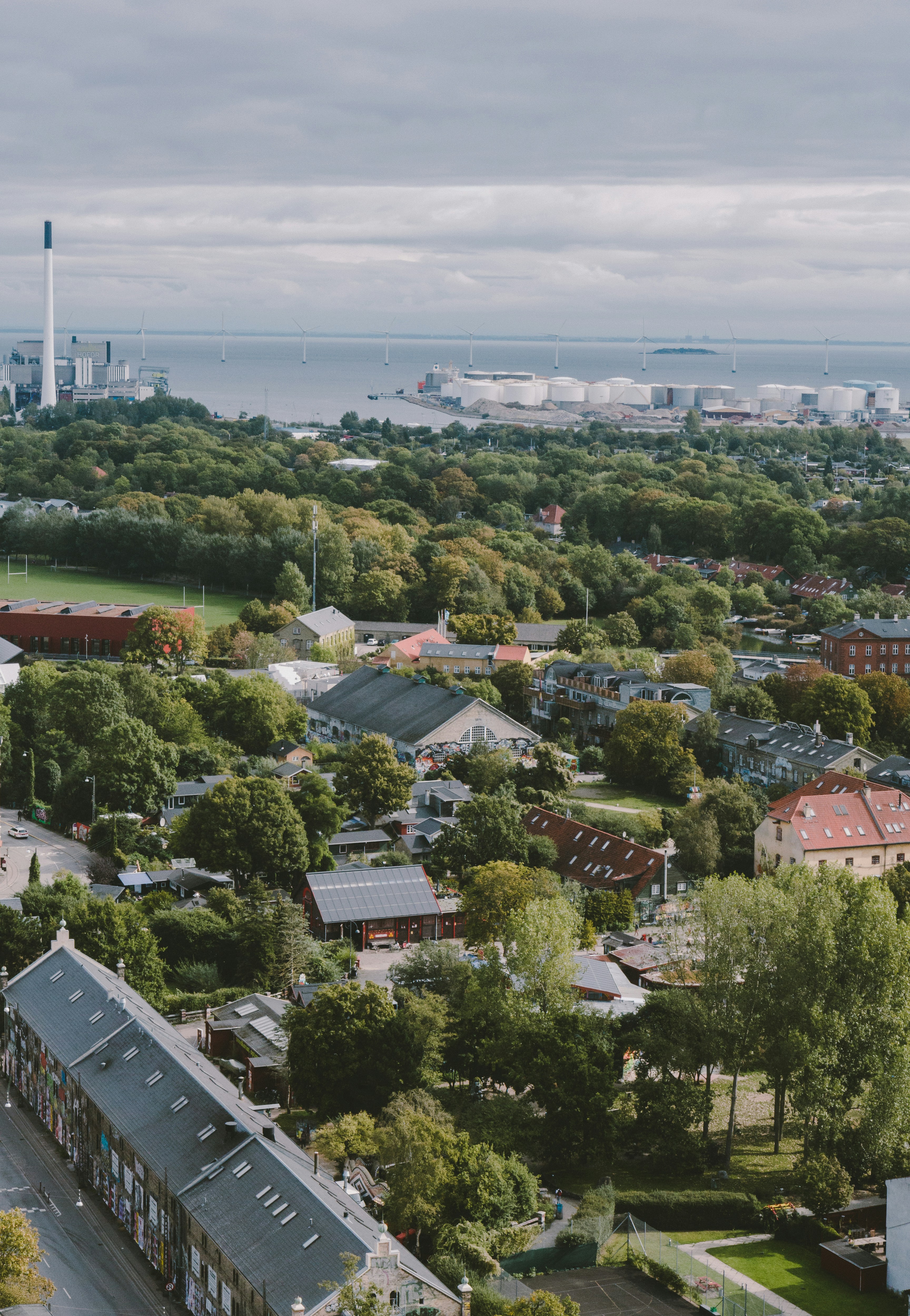Aerial view of Christiania's lush greenery and colorful rooftops, with industrial structures and wind turbines along the coastline.