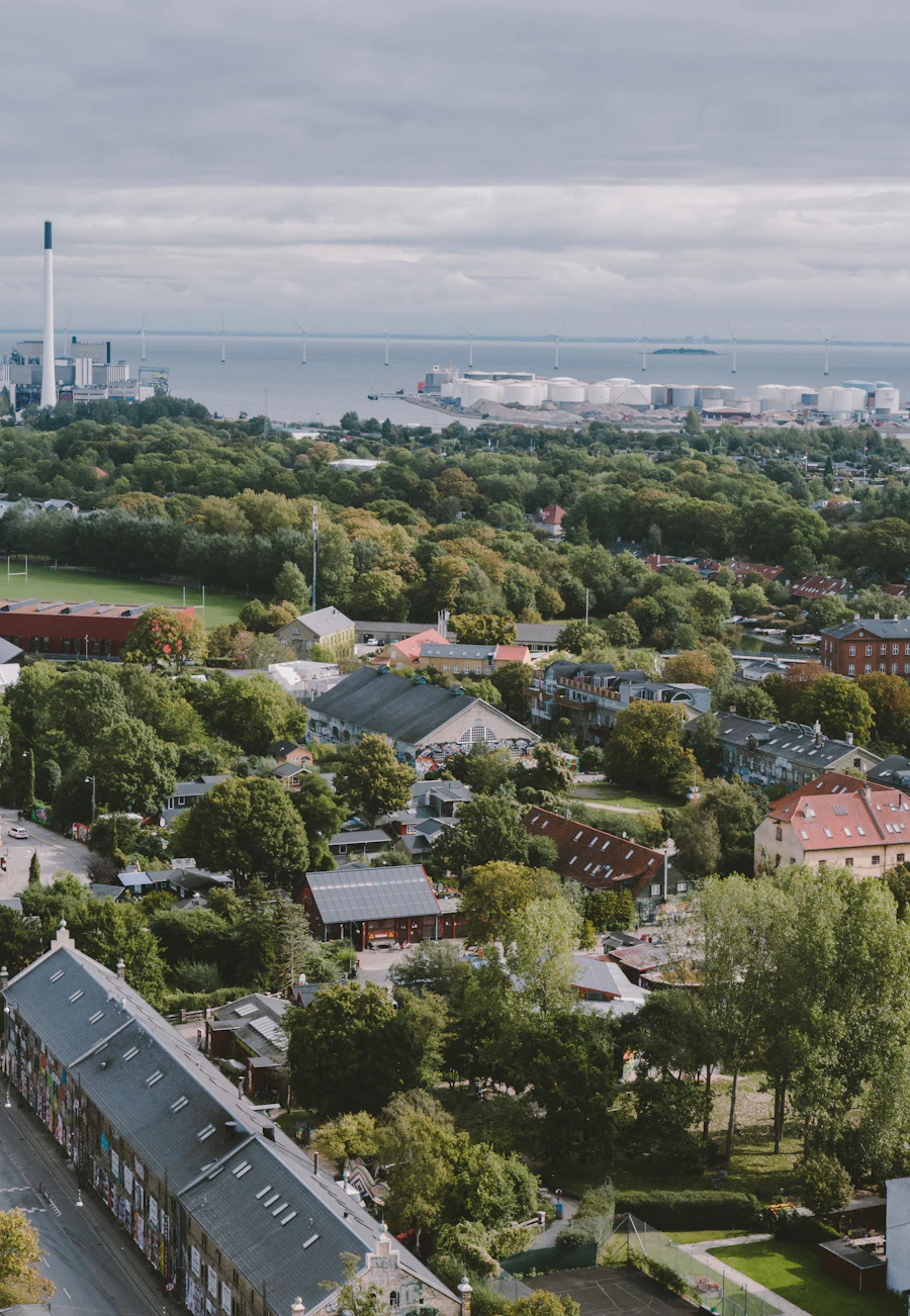 Elevated view over a leafy residential district toward an industrial harbor, with water, storage tanks, and wind turbines on the horizon. A tall smokestack rises at the left under an overcast sky.