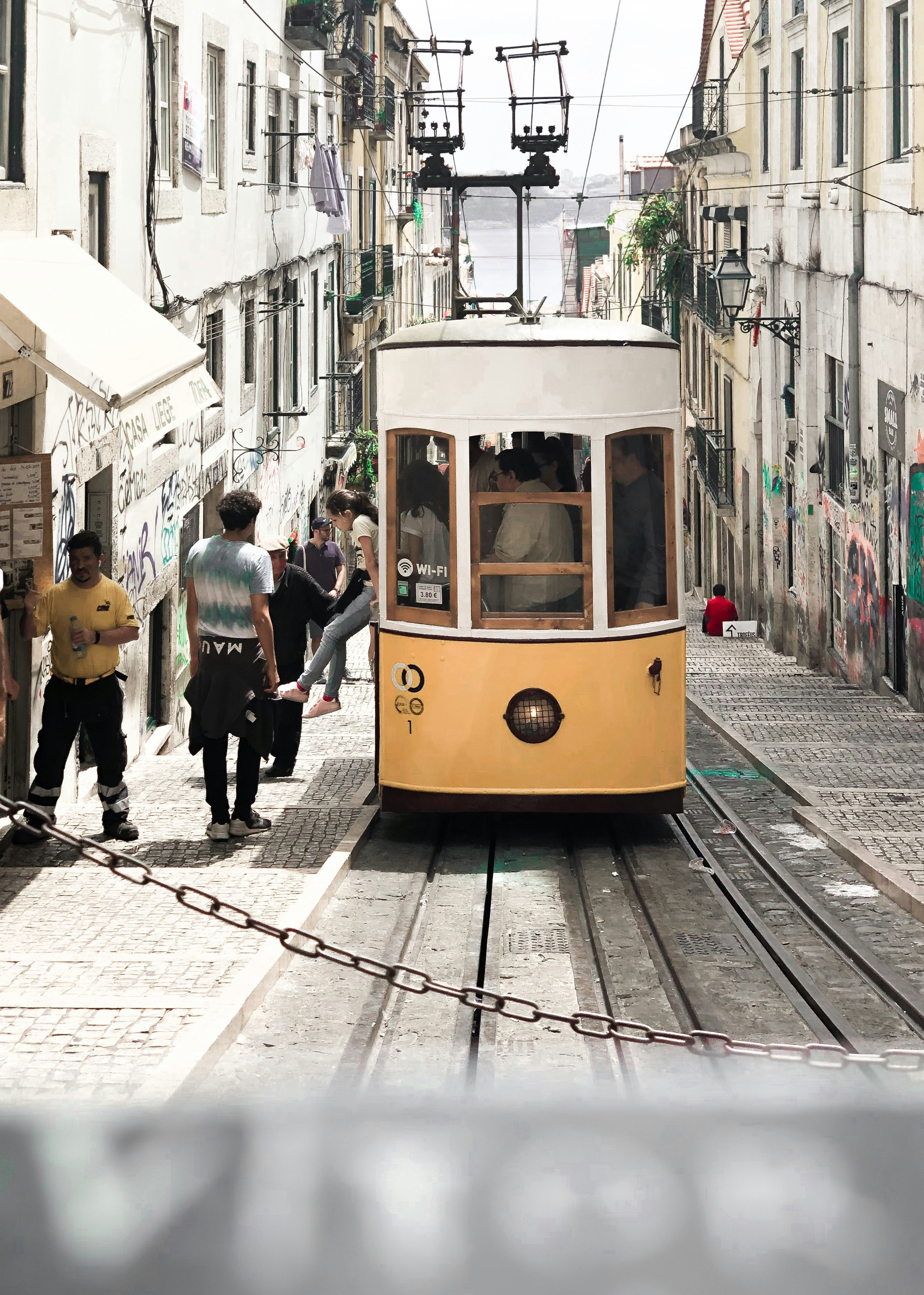 A vintage tram navigates a narrow, graffiti-adorned street in Lisbon, surrounded by pedestrians and vibrant city life.
