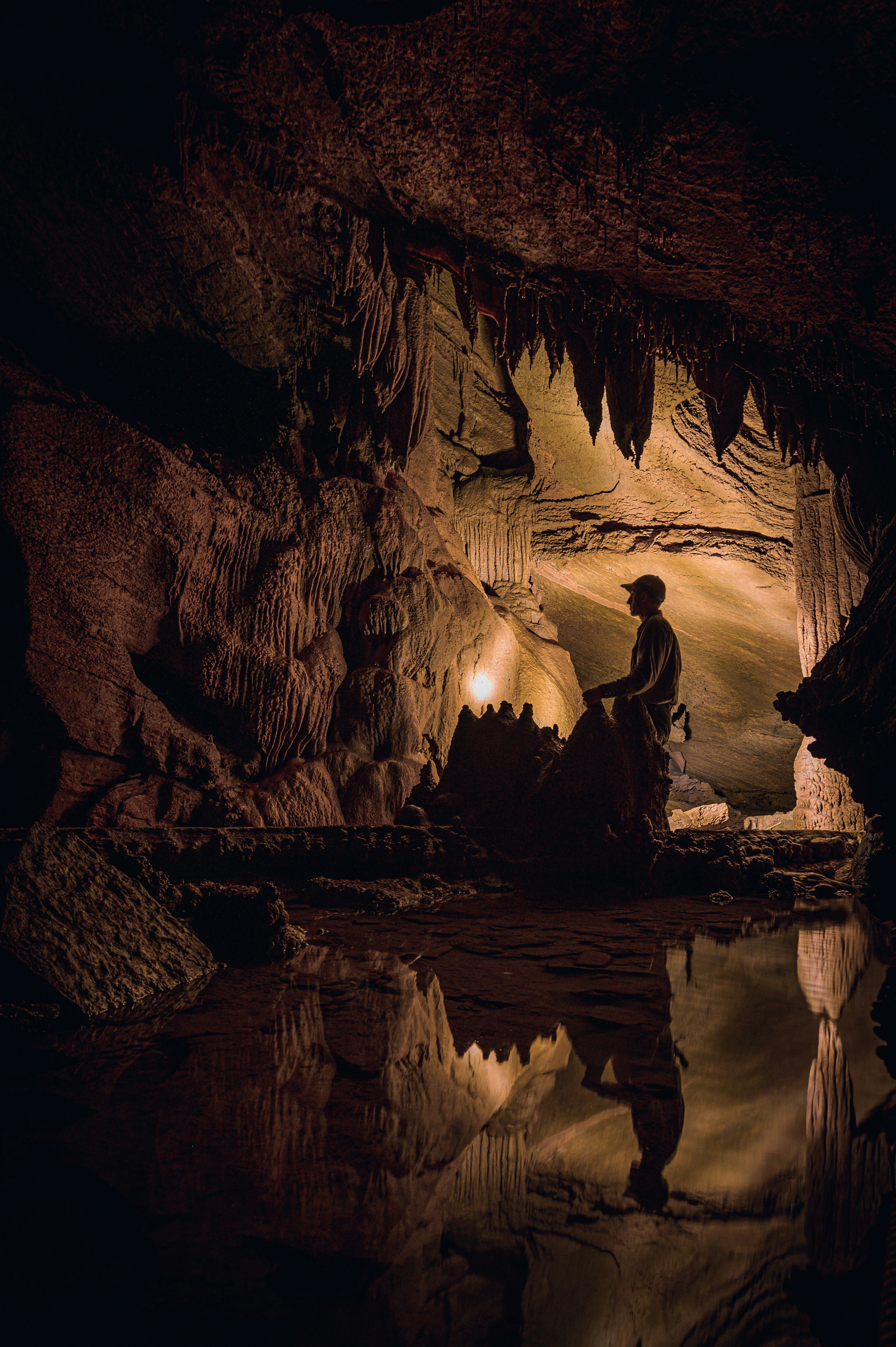 A solitary figure stands in a cavern, illuminated by soft light, reflecting on the still water below. Intricate rock formations create a striking backdrop.