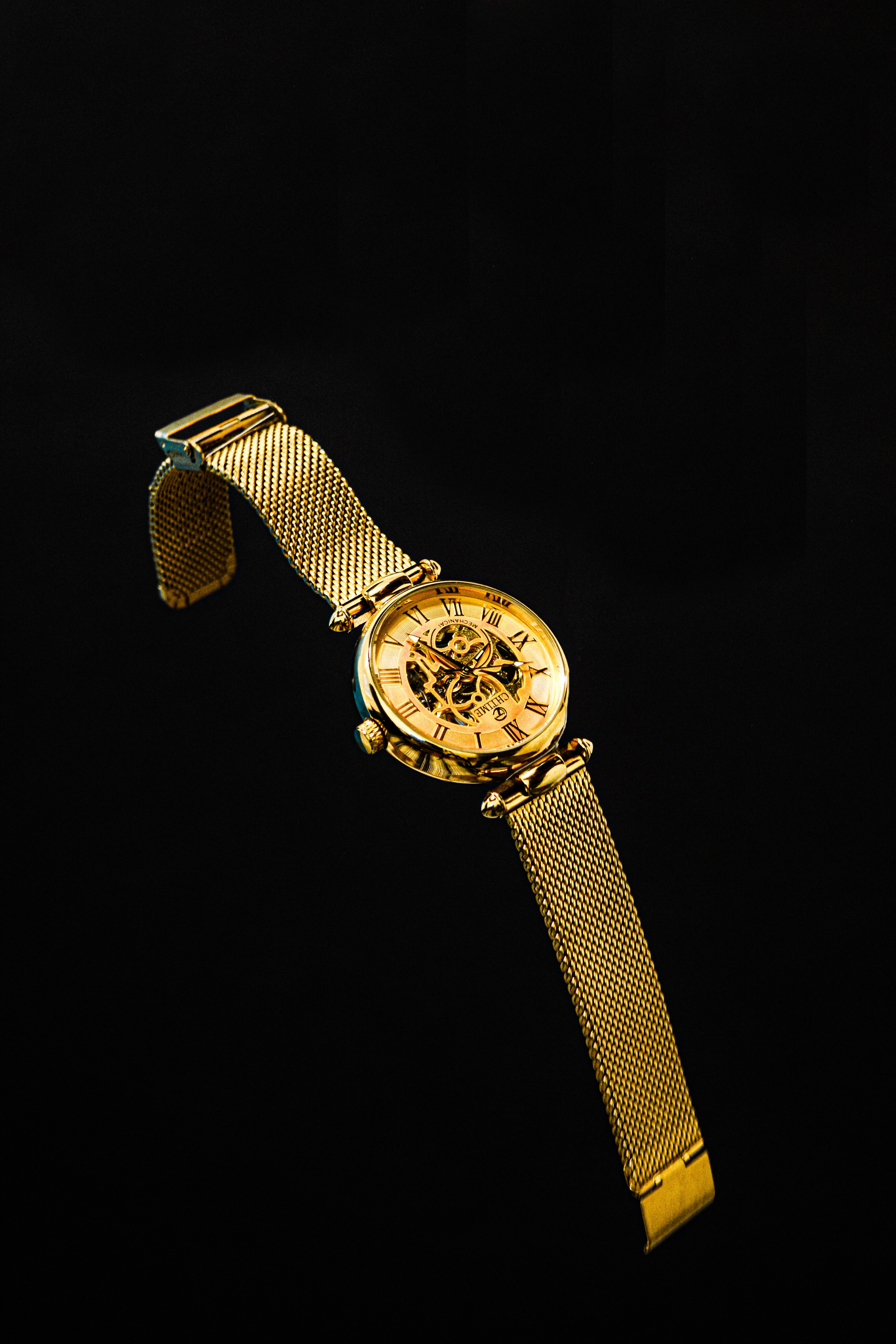 Gold watch with intricate dial and mesh strap, reflecting light against a dark backdrop.