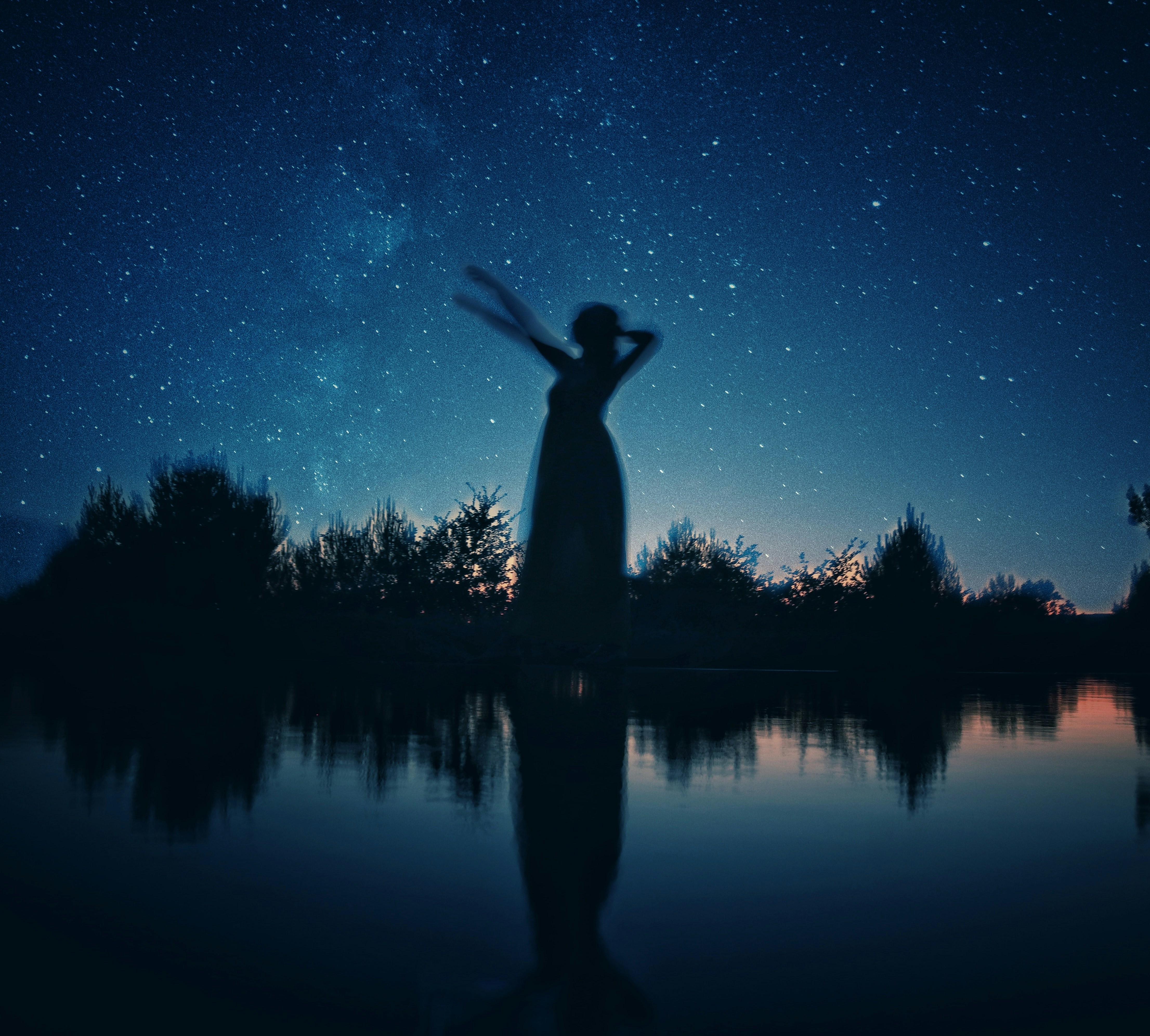 Silhouette of a woman standing by a pool under a starry night sky with her reflection in the water.