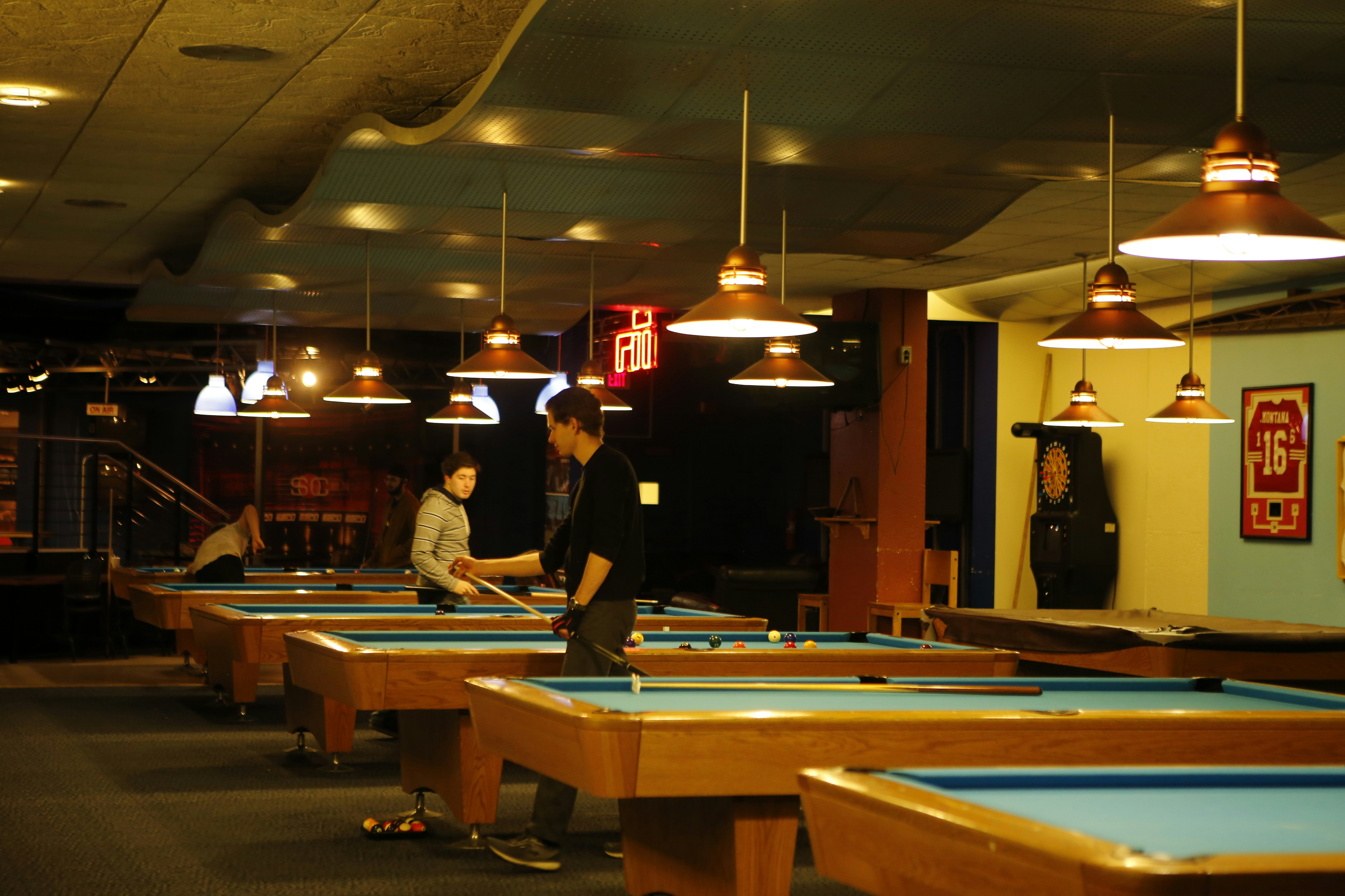 Two players engaged in a game of pool in a dimly lit recreation area, surrounded by several pool tables and warm overhead lights.