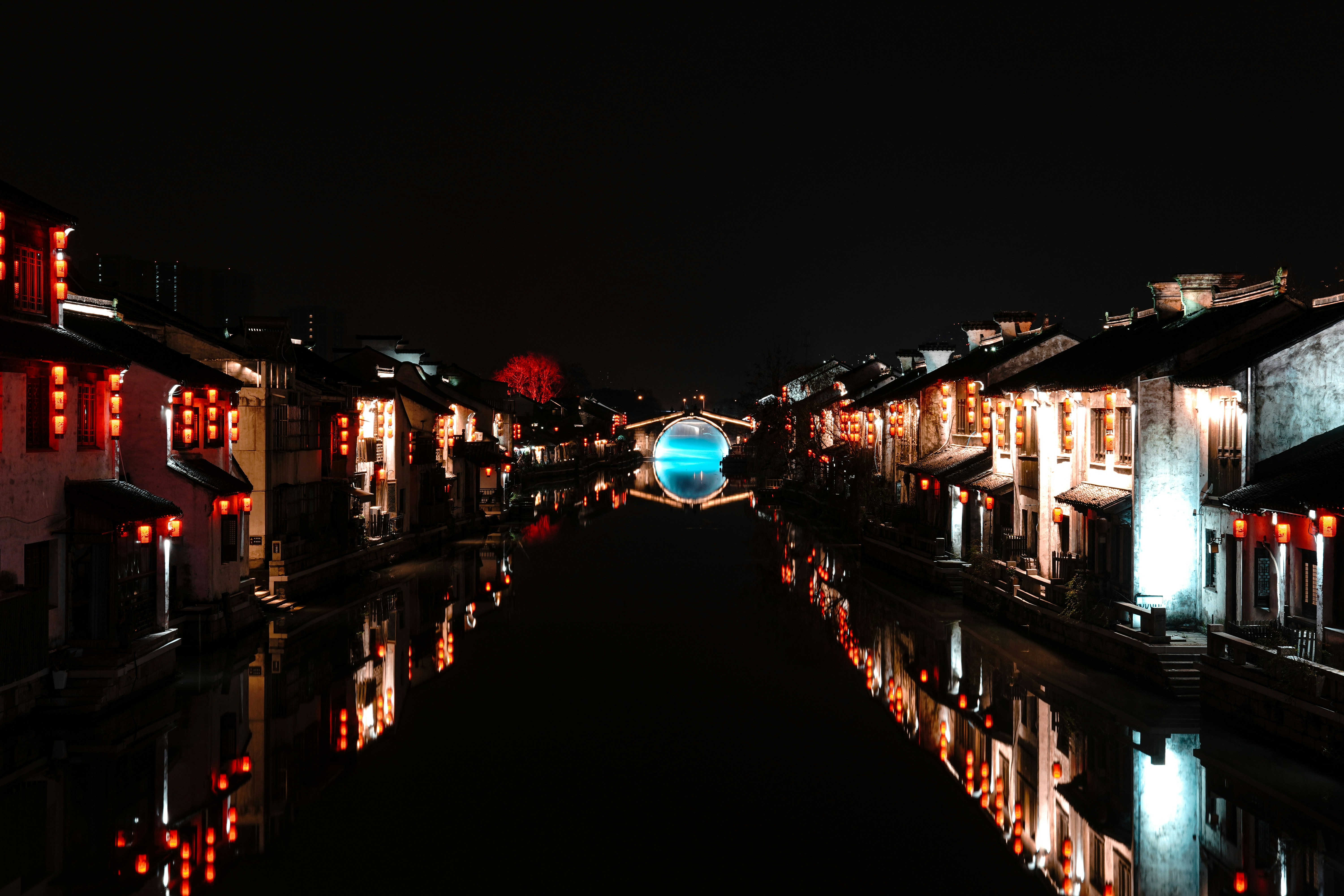 Traditional buildings with red lanterns reflecting on a calm canal at night.