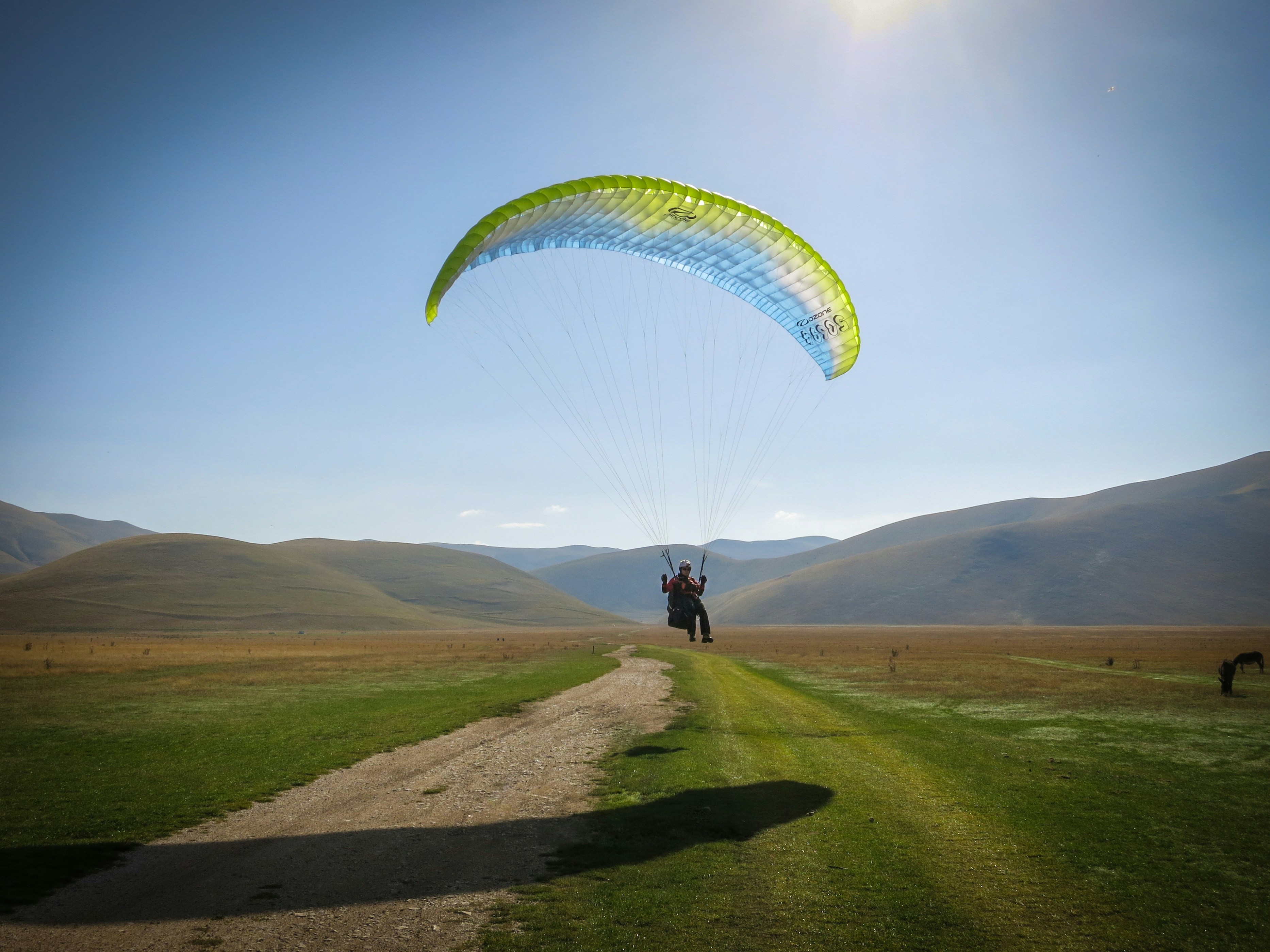 Paraglider glides above a grassy path through a rolling valley under a bright blue sky.