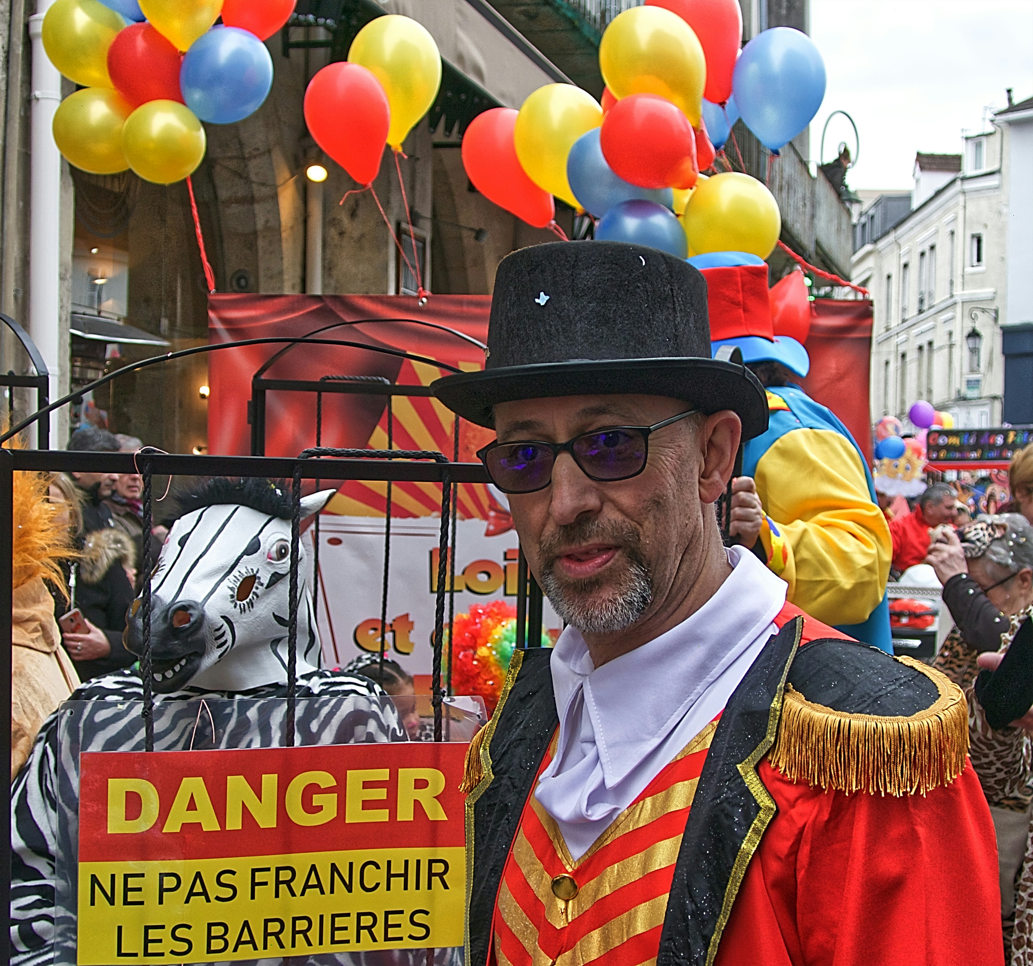 A performer in a vibrant costume stands beside a zebra mascot, with colorful balloons floating in the background. A caution sign adds a humorous touch to the festive atmosphere.