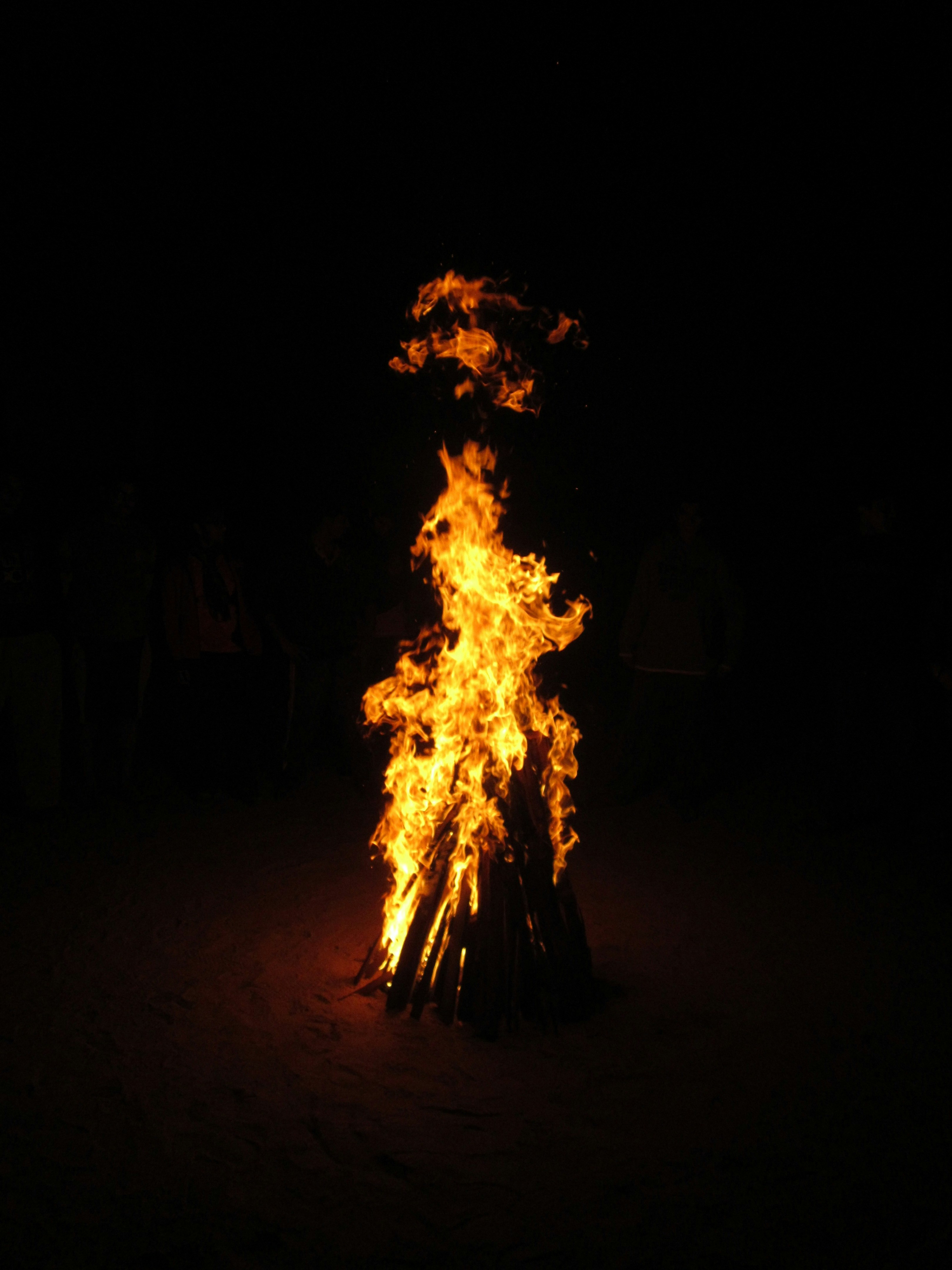 Night photograph of a tall bonfire built from stacked logs, with flames leaping upward and casting warm light on surrounding silhouettes. The composition centers the blaze to emphasize a communal gathering around the fire.