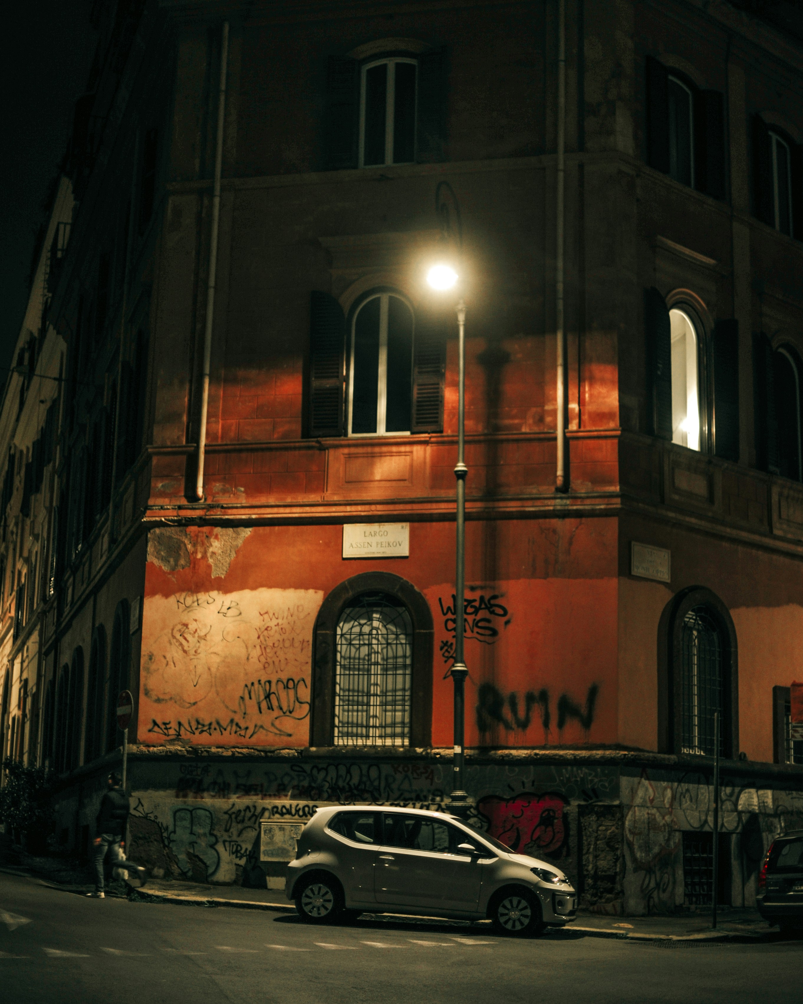 Illuminated corner of a weathered building adorned with graffiti, showcasing urban art against a backdrop of night. A parked car adds to the scene's ambiance.