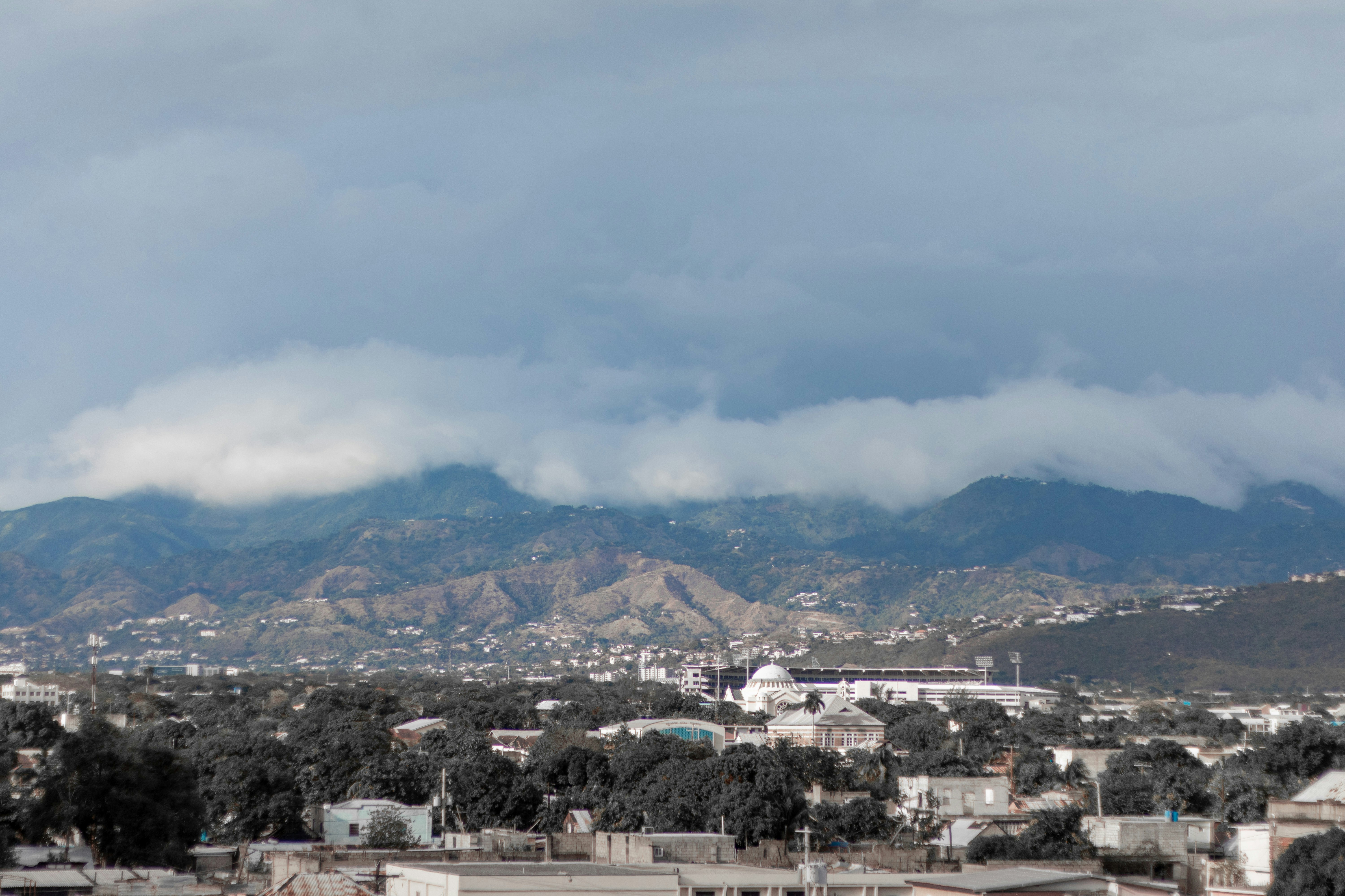 Cityscape with buildings sprawling towards distant mountains under a cloudy sky.